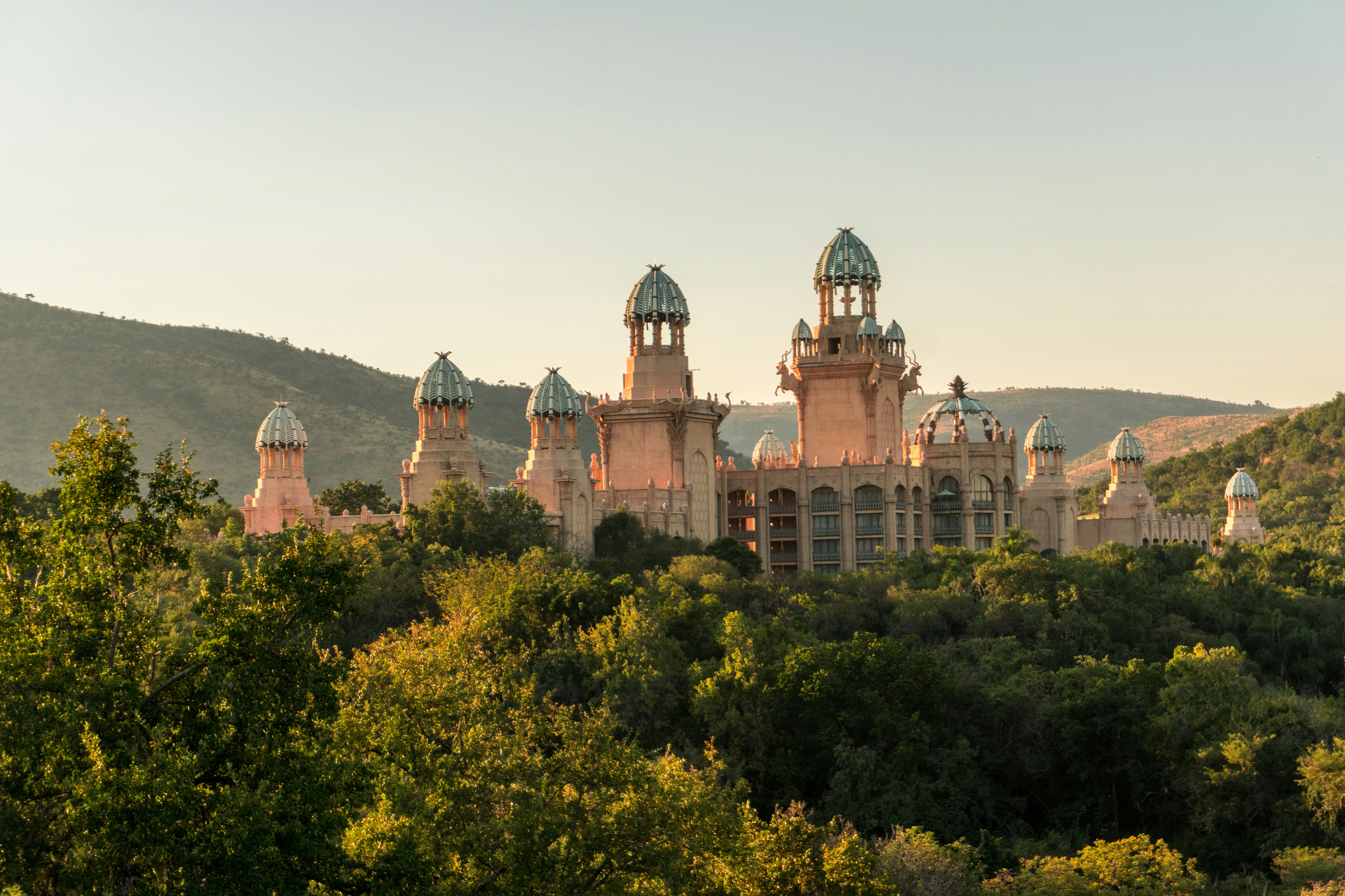 Grand palace with domed towers nestled among lush greenery and rolling hills under a clear sky.