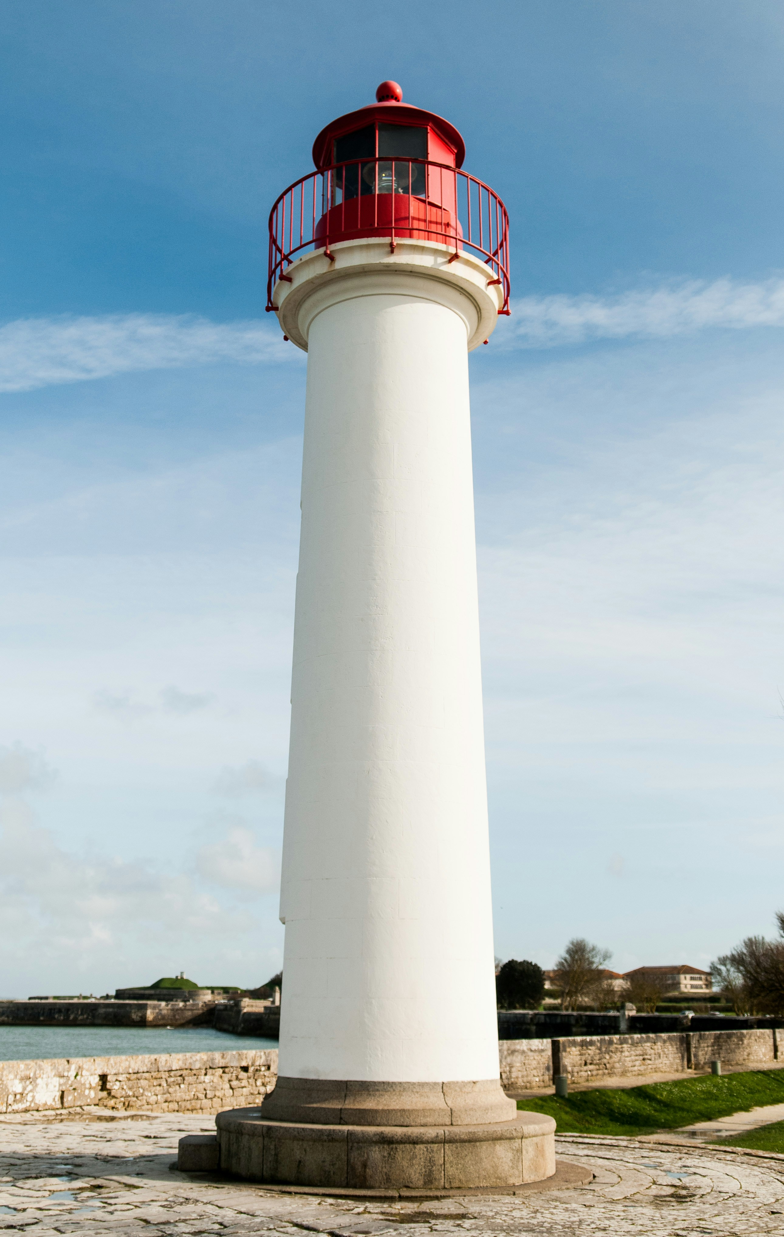A tall, white lighthouse with a red top stands against a blue sky, overlooking a coastal landscape. It symbolizes maritime navigation and local history.