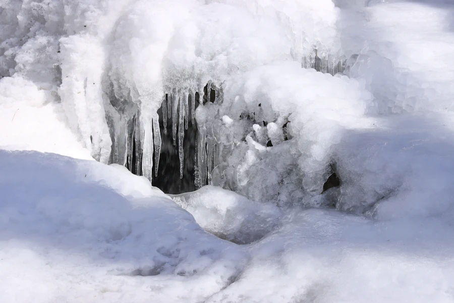 Snow-covered rocks with a dark hollow at center, lined by hanging icicles and frosted ice. Soft winter light highlights the textured snow and frozen surfaces.