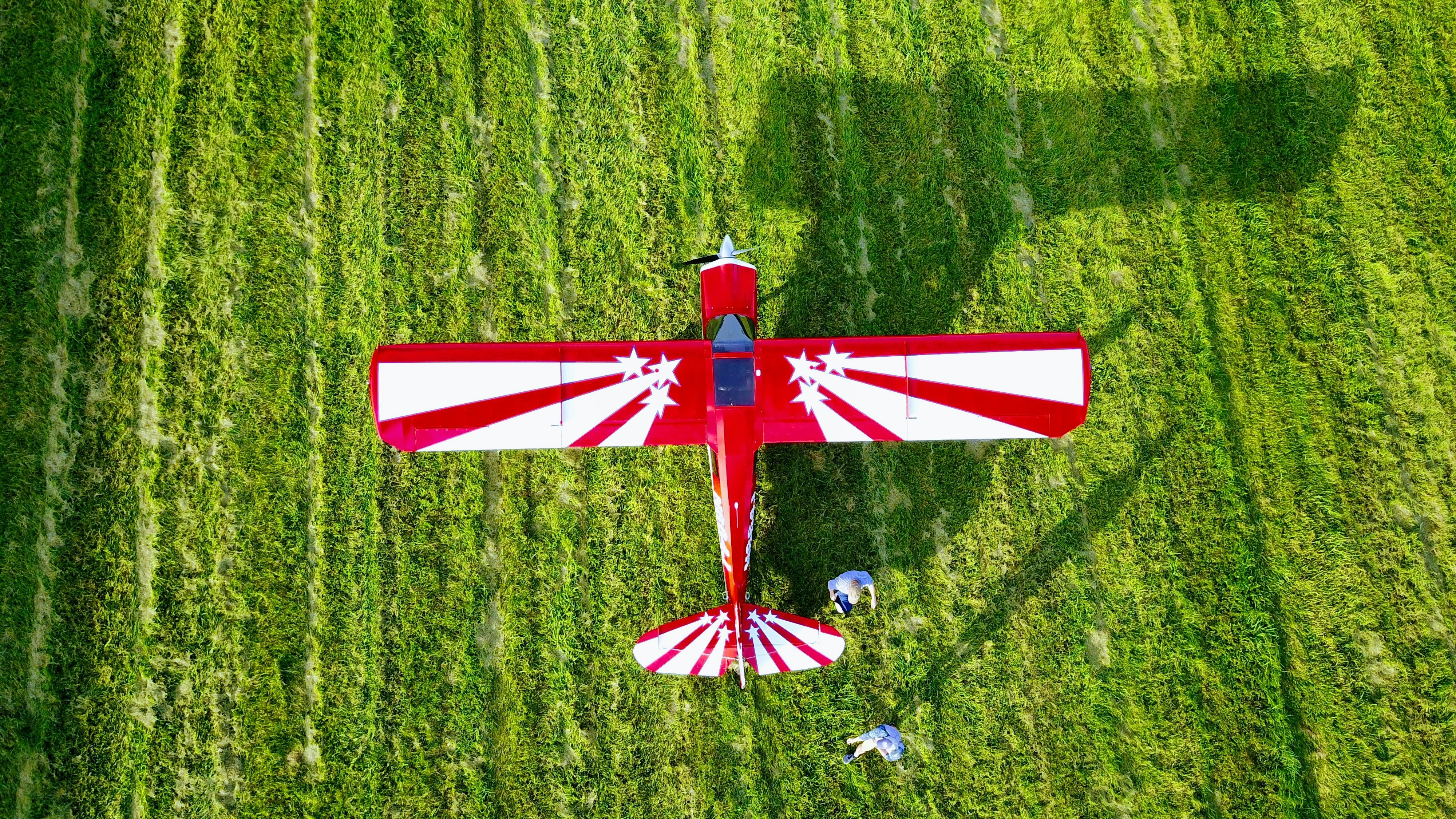 A vibrant red and white stunt plane rests on a grassy field, with two individuals inspecting it. The overhead perspective highlights the plane's striking design against the lush green backdrop.