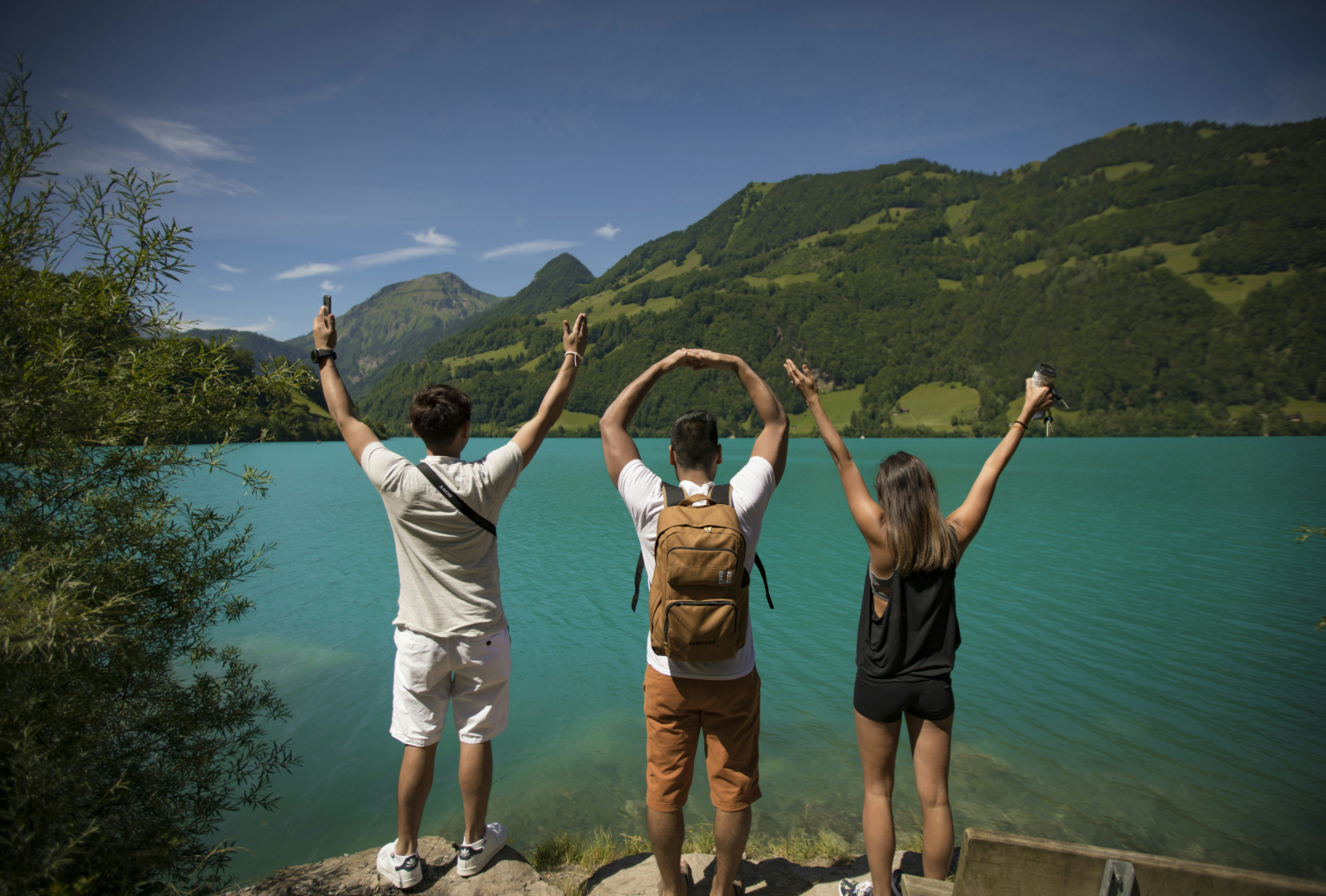Three friends joyfully celebrating at the edge of a vibrant turquoise lake, surrounded by lush green mountains.