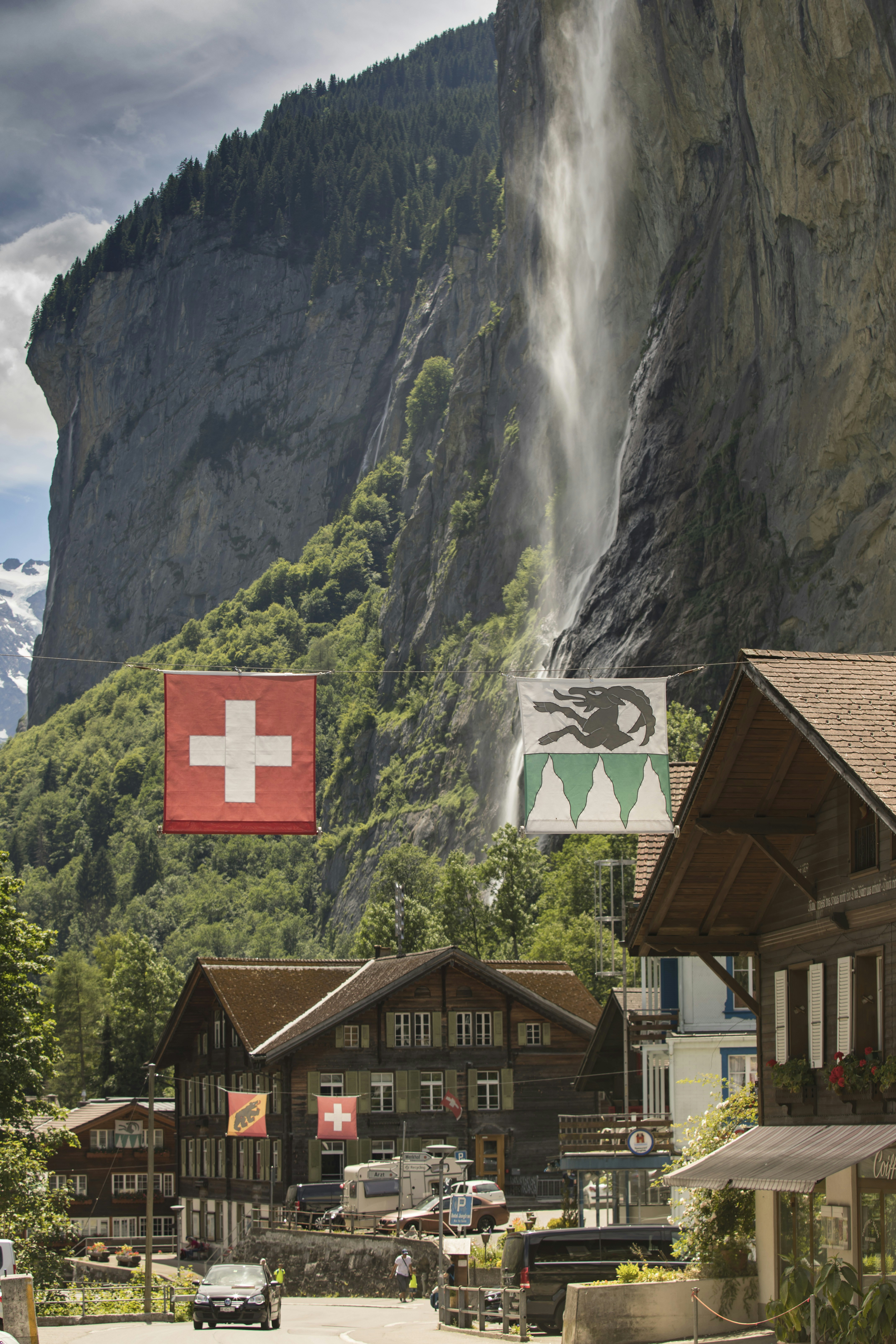 Charming Swiss village with traditional wooden houses and vibrant flags, framed by towering mountains and a cascading waterfall in the background.