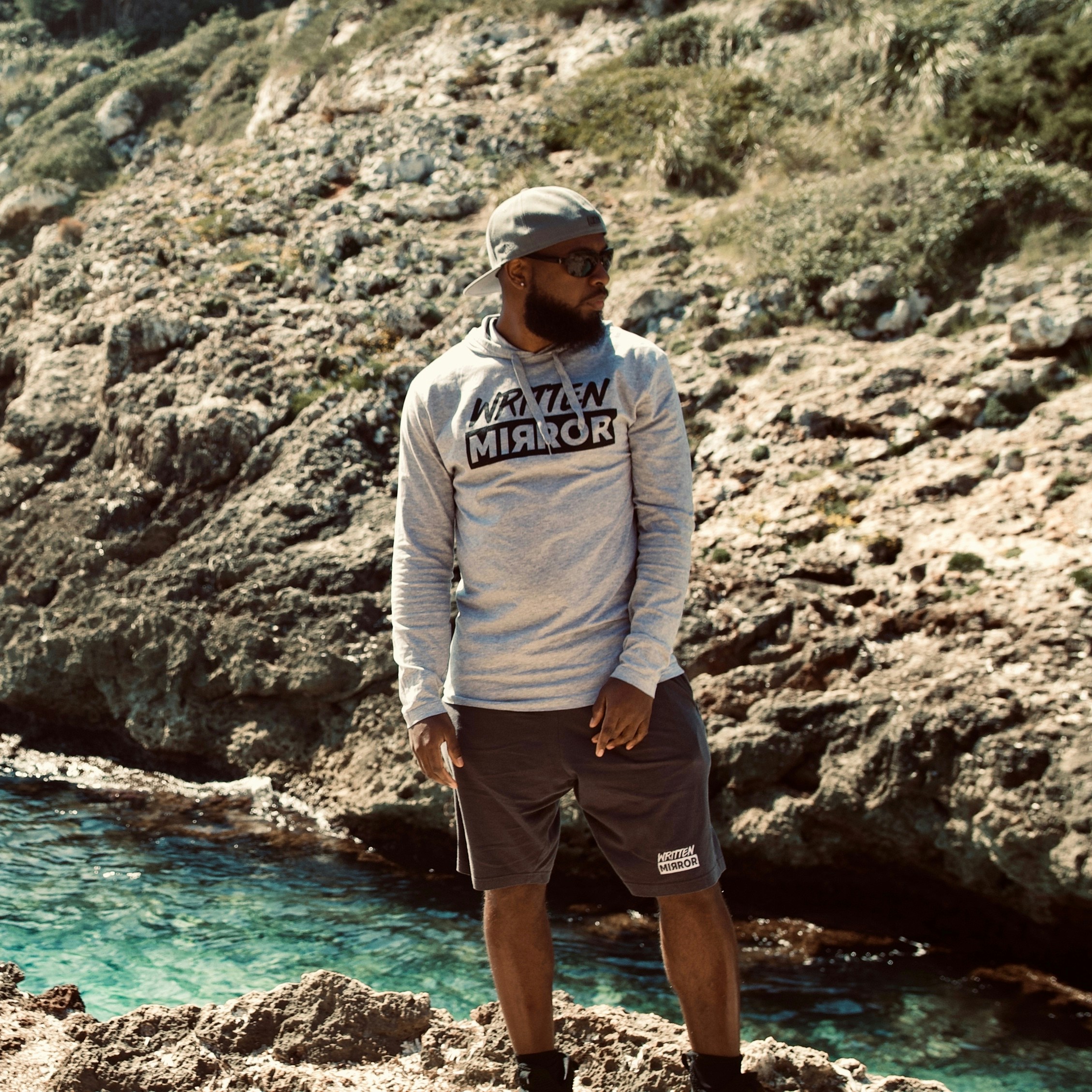 Individual in casual attire stands thoughtfully near rocky coastline, with clear turquoise water in the background.