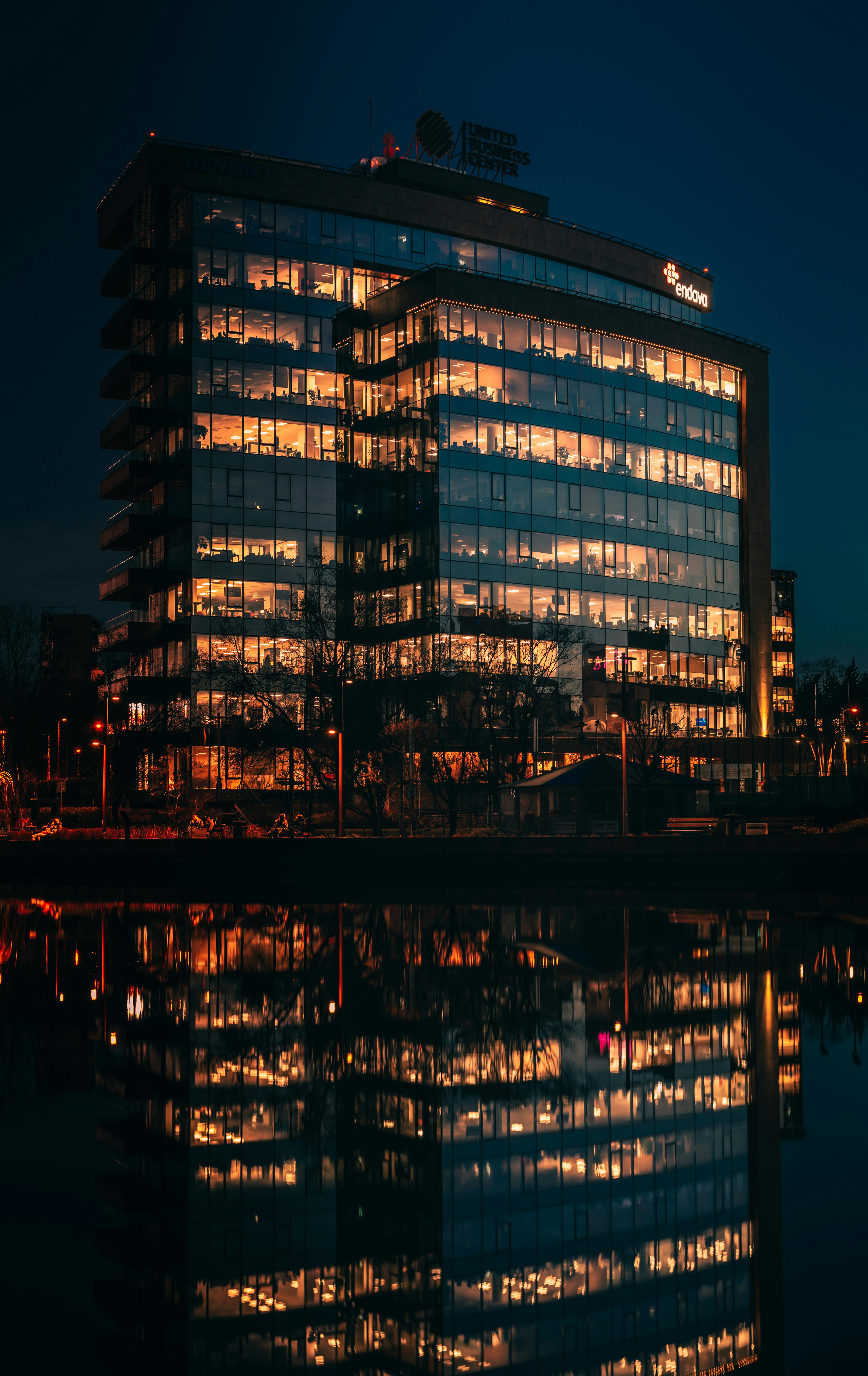 Modern office building illuminated at night, reflecting in the still water below. The glow from the windows creates a warm ambiance.