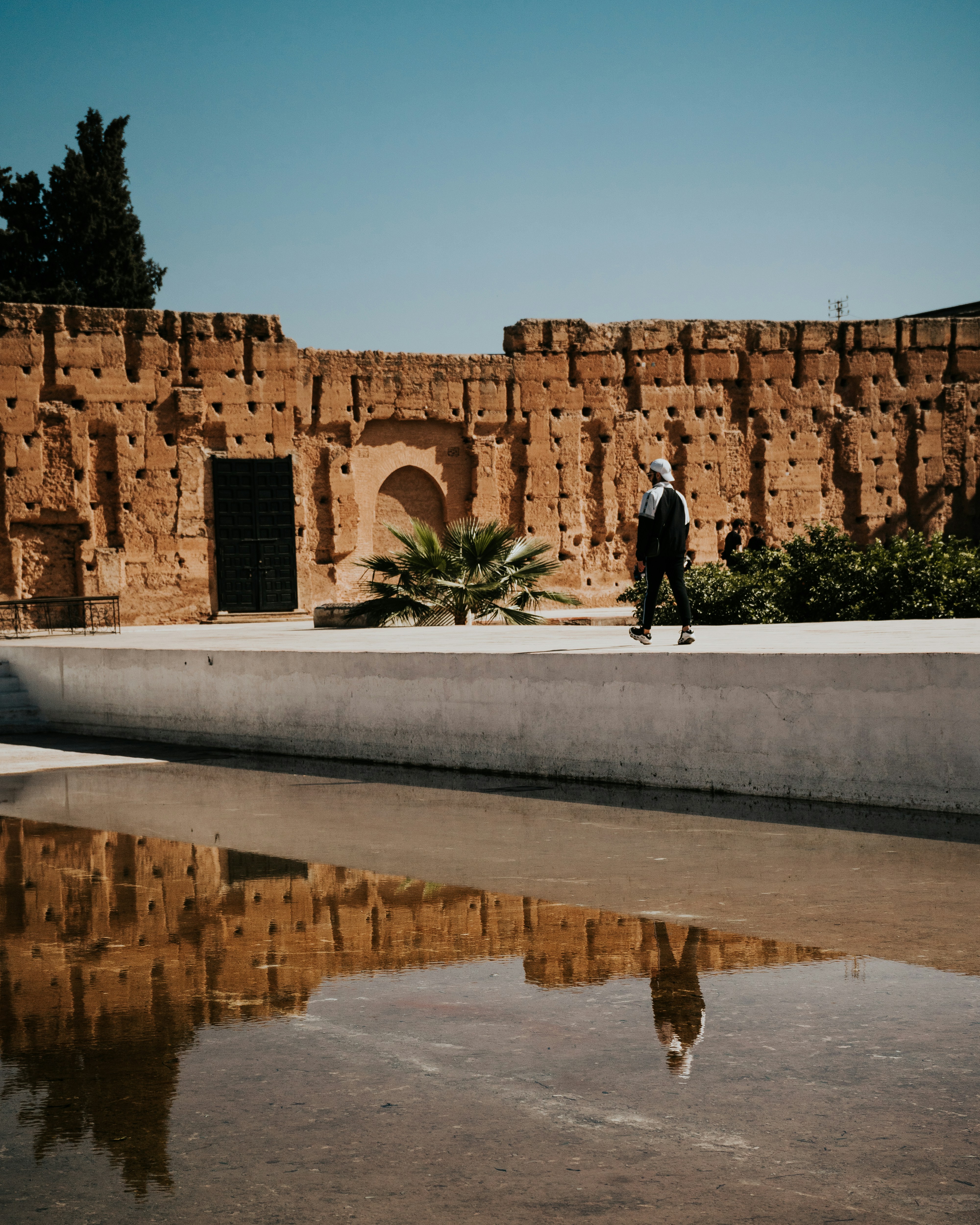A person walks along a reflective pathway beside ancient walls and lush greenery, capturing a moment of tranquility amidst historical architecture.