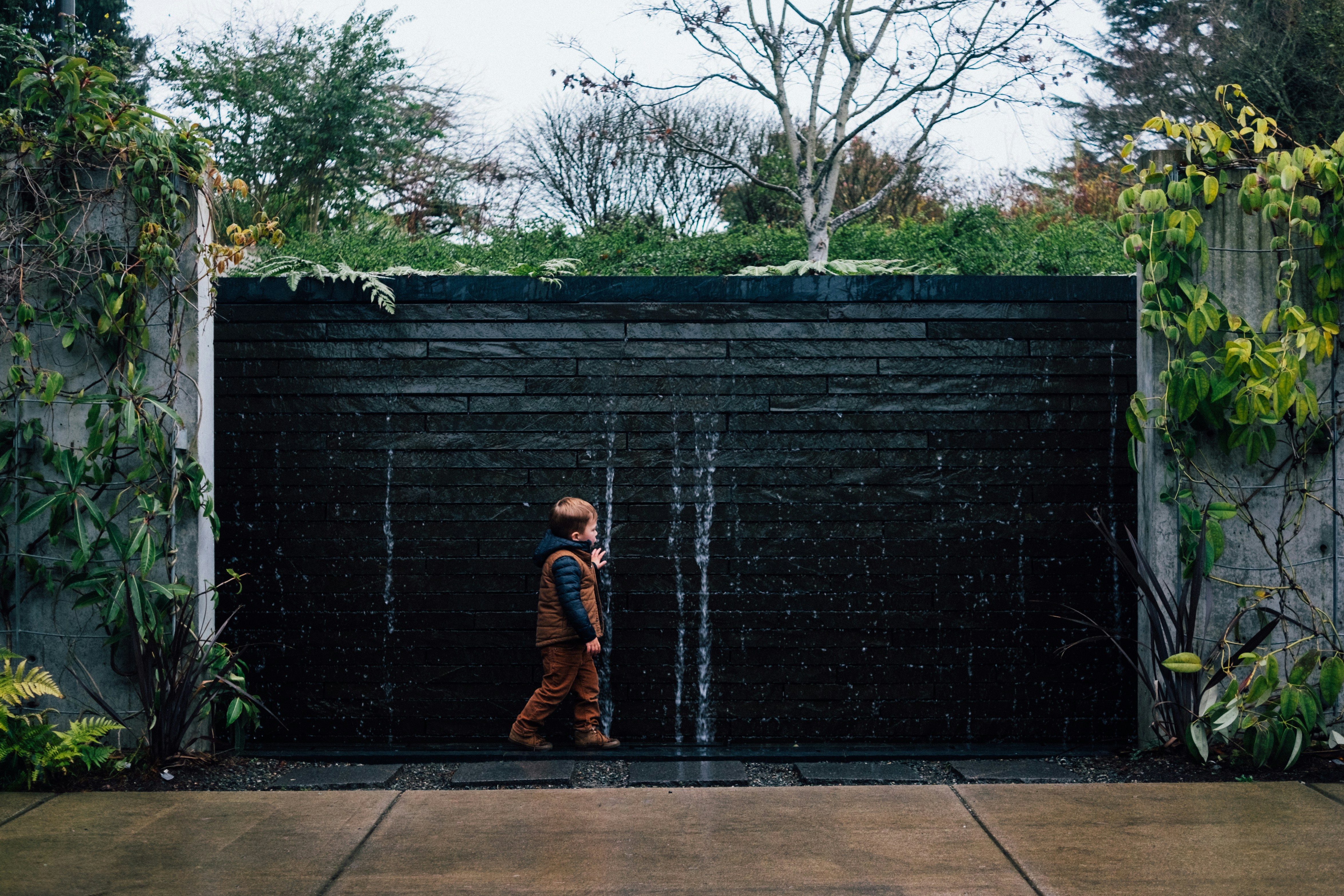 Person in brown clothing walking past a dark, textured waterfall wall surrounded by greenery.