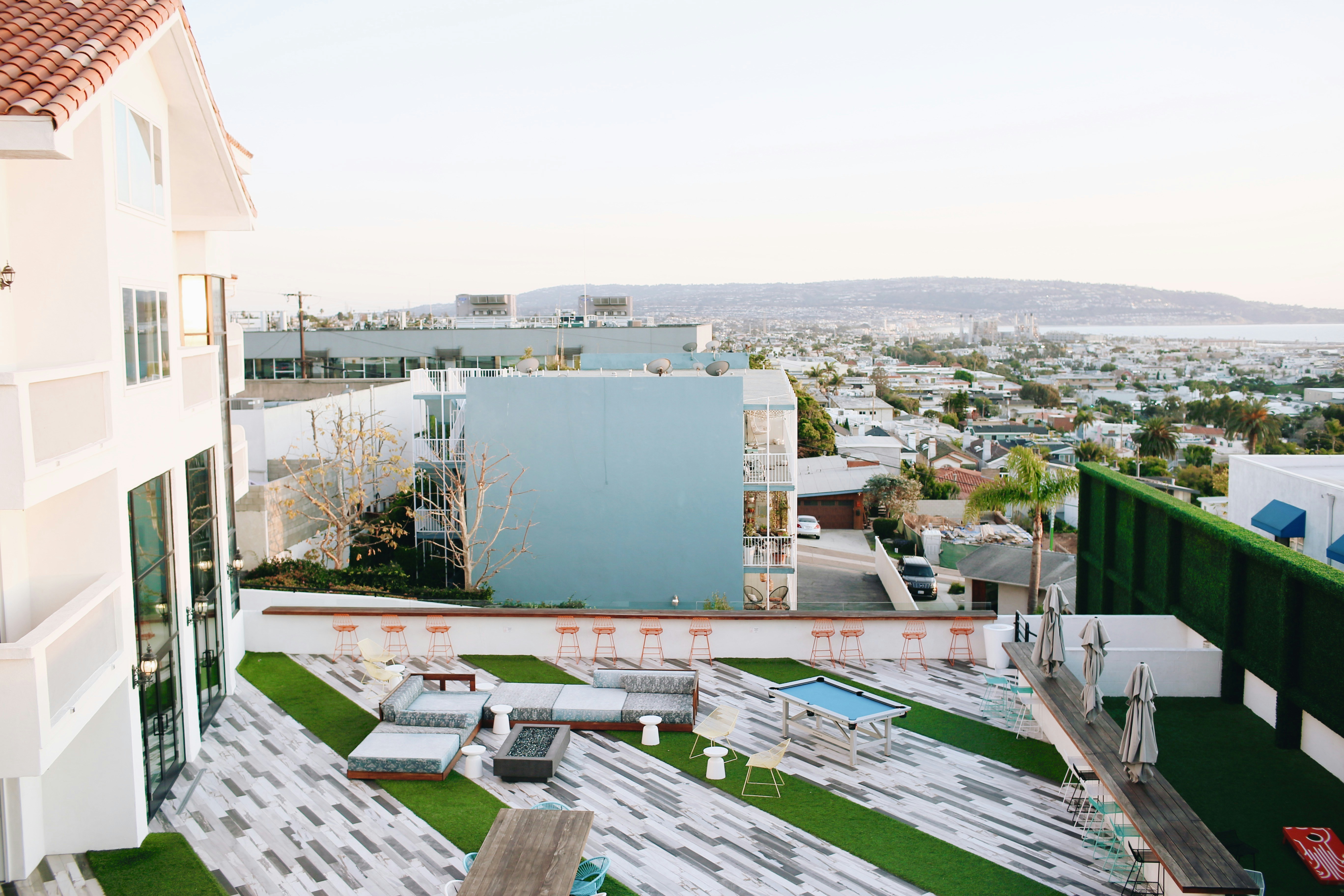 Sunlit rooftop pool terrace with modern lounge areas overlooking a coastal cityscape and distant hills.