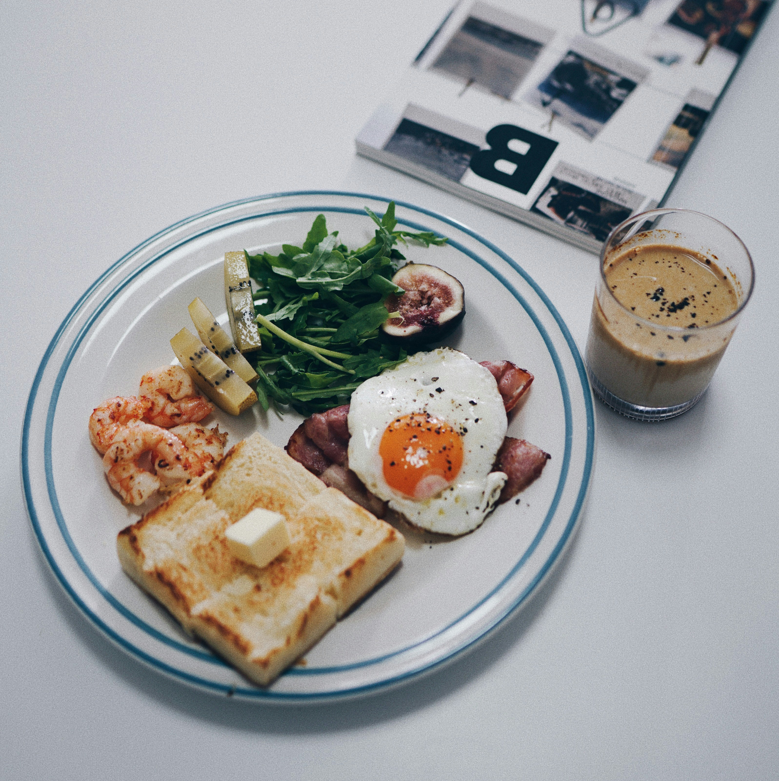 A beautifully arranged breakfast plate featuring fried egg, bacon, shrimp, toast with butter, and a side of greens and figs, accompanied by a glass of coffee.