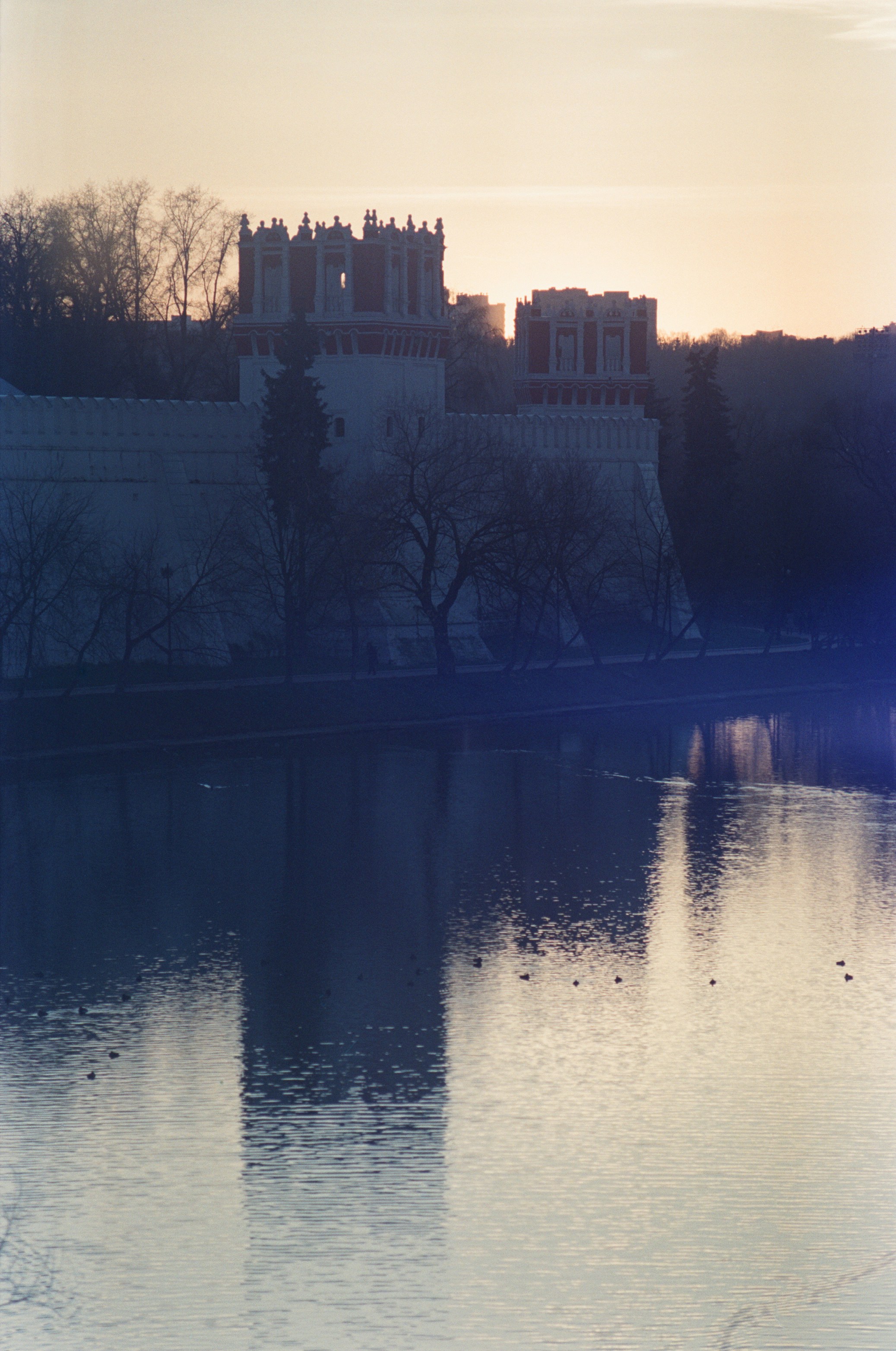 Silhouetted towers and trees reflect in a calm river at sunset, with a peaceful sky overhead.