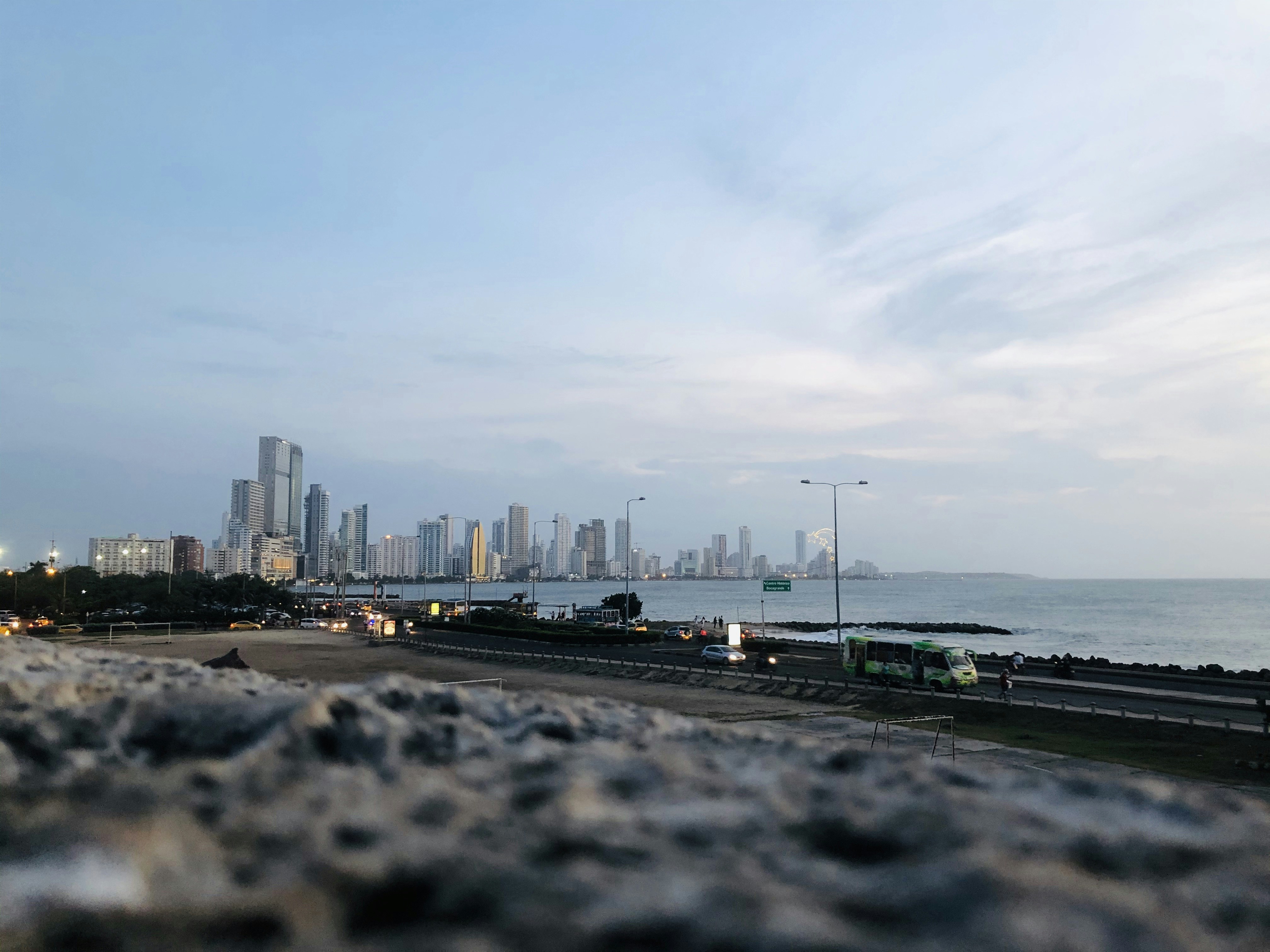 City skyline with skyscrapers under a partly cloudy sky, viewed from a rocky shoreline during daytime.