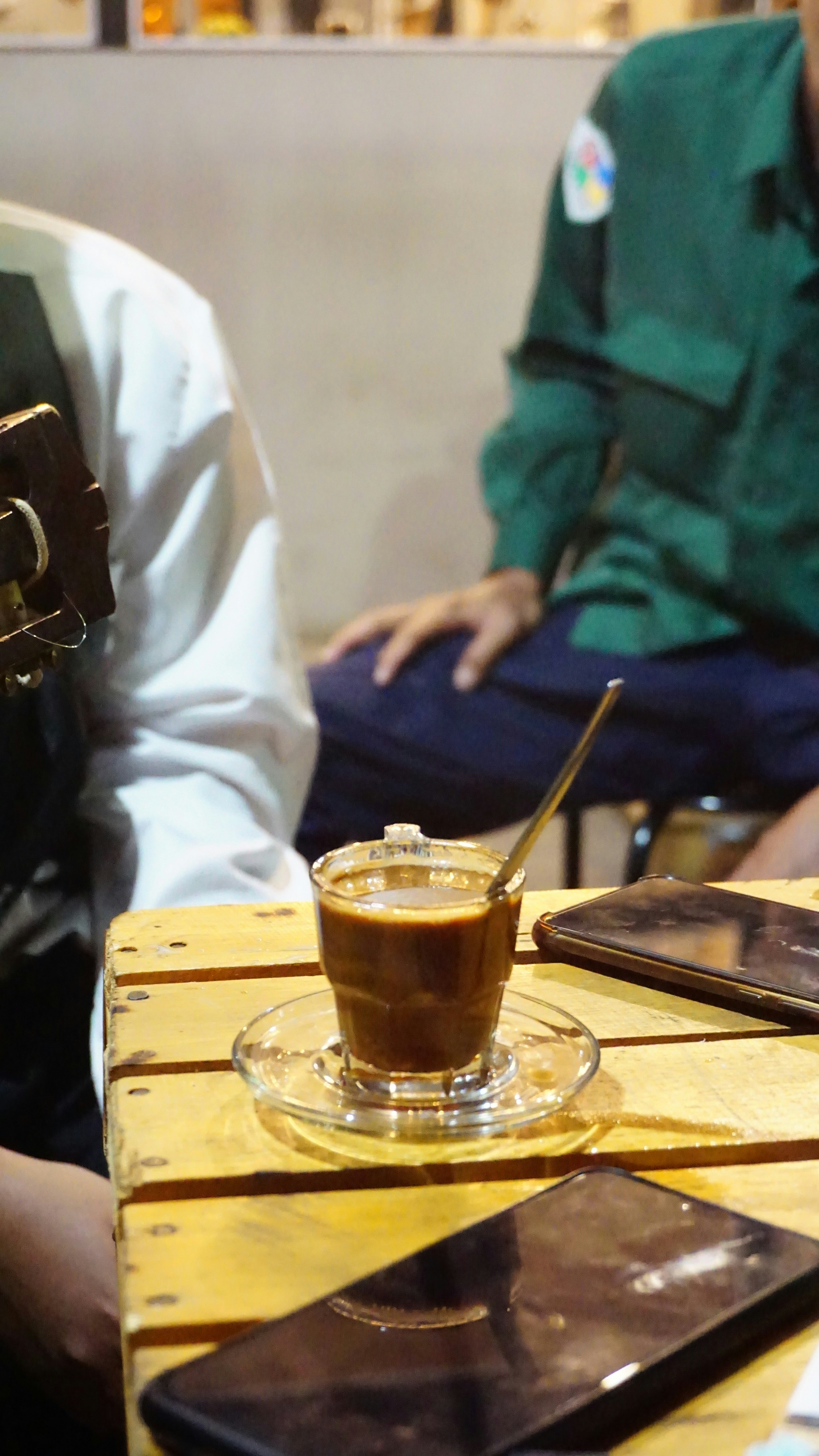 A small glass of coffee sits on a wooden table surrounded by mobile phones, hinting at a casual gathering. The atmosphere suggests a blend of conversation and relaxation.