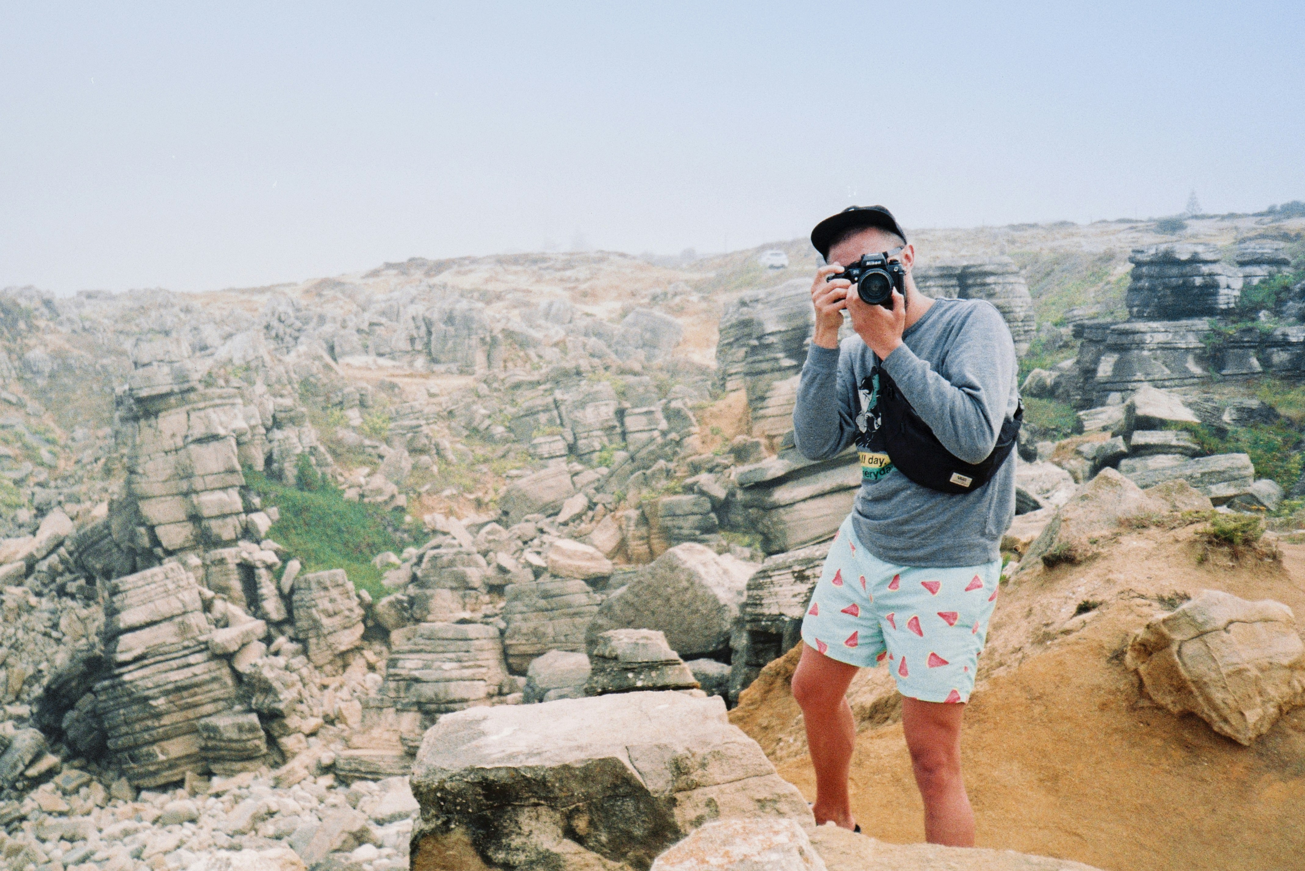 A photographer standing on a rocky outcrop captures the rugged landscape, showcasing unique geological formations under a hazy sky.