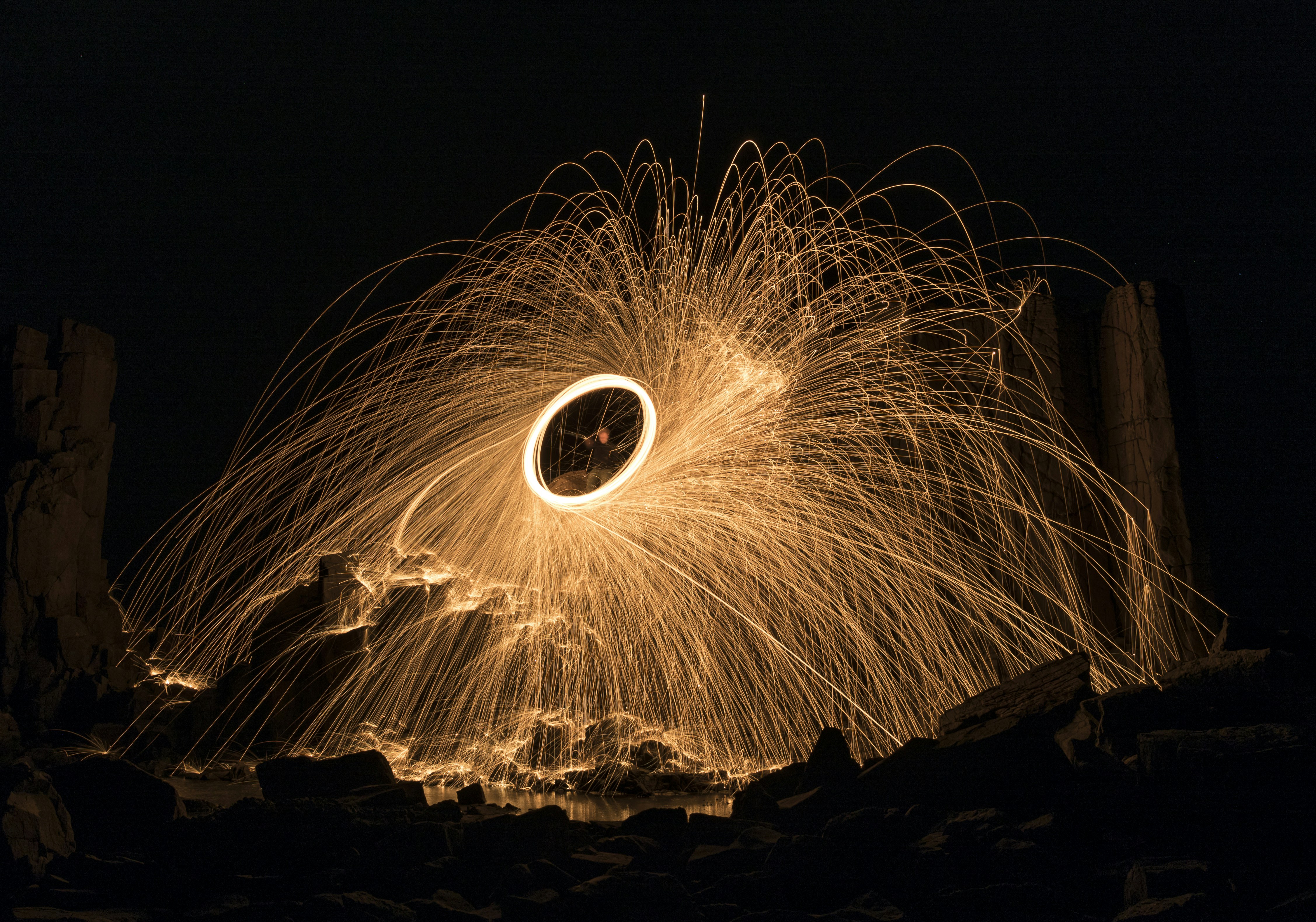 Long exposure capturing sparks flying from a central point amidst rocky terrain at night. The dynamic flow of light creates an enchanting visual spectacle.