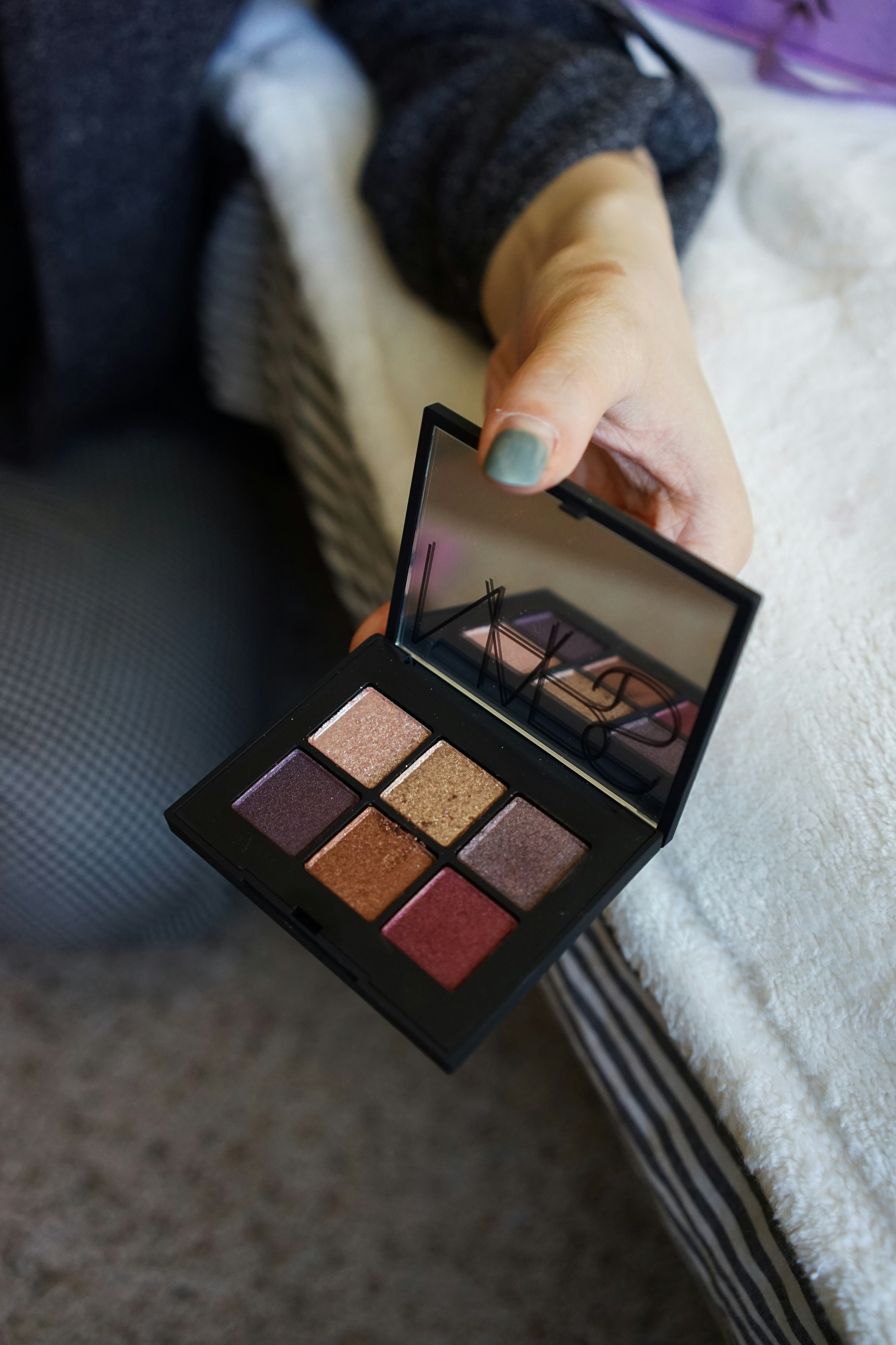 A hand showcasing an open makeup palette featuring a variety of eyeshadow colors, including shimmering and matte shades. The setting suggests a casual beauty routine.