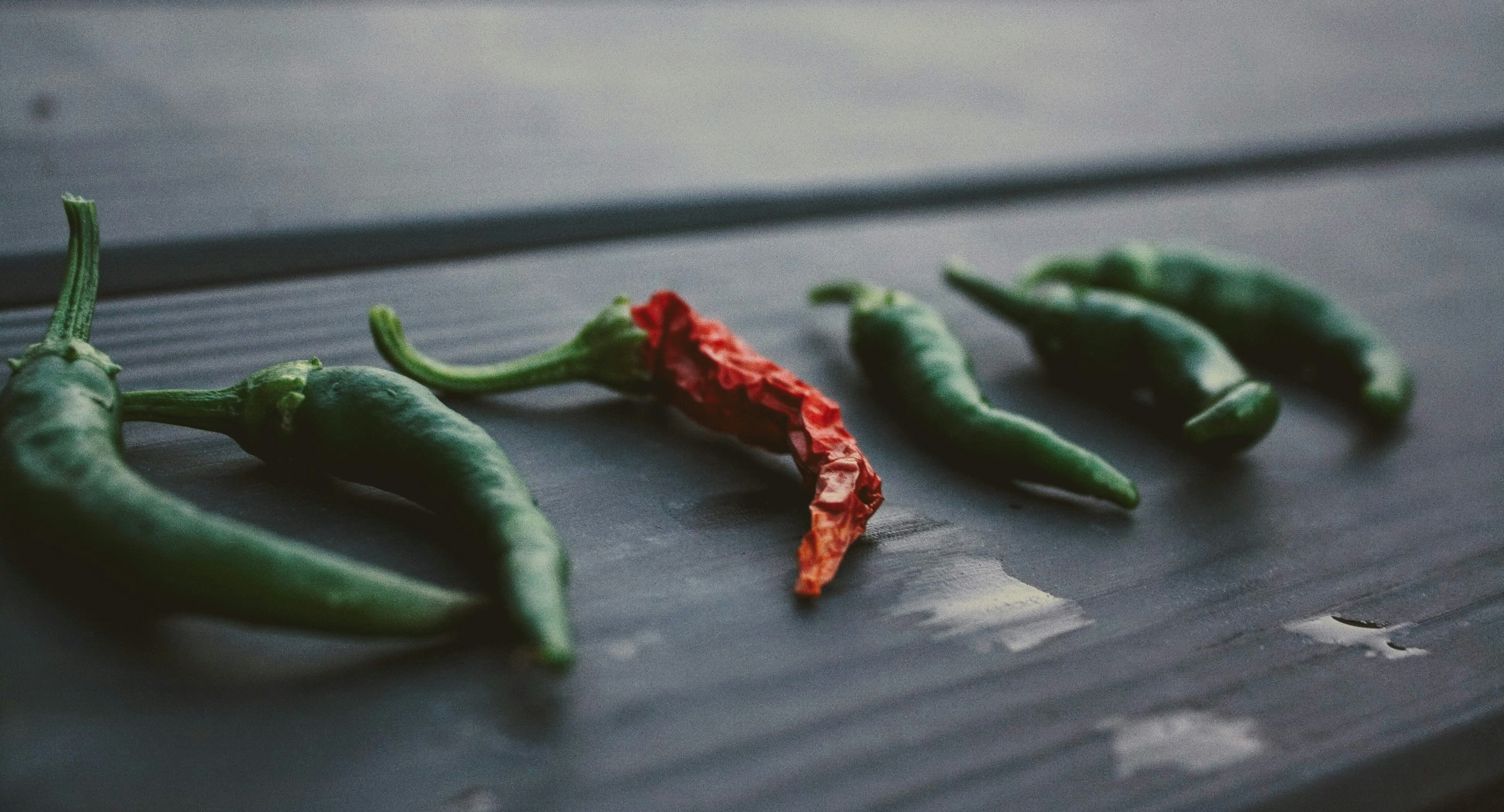 Five chili peppers arranged in a row on a wooden surface, showcasing their varying colors and textures. The vibrant red pepper stands out among the green ones.