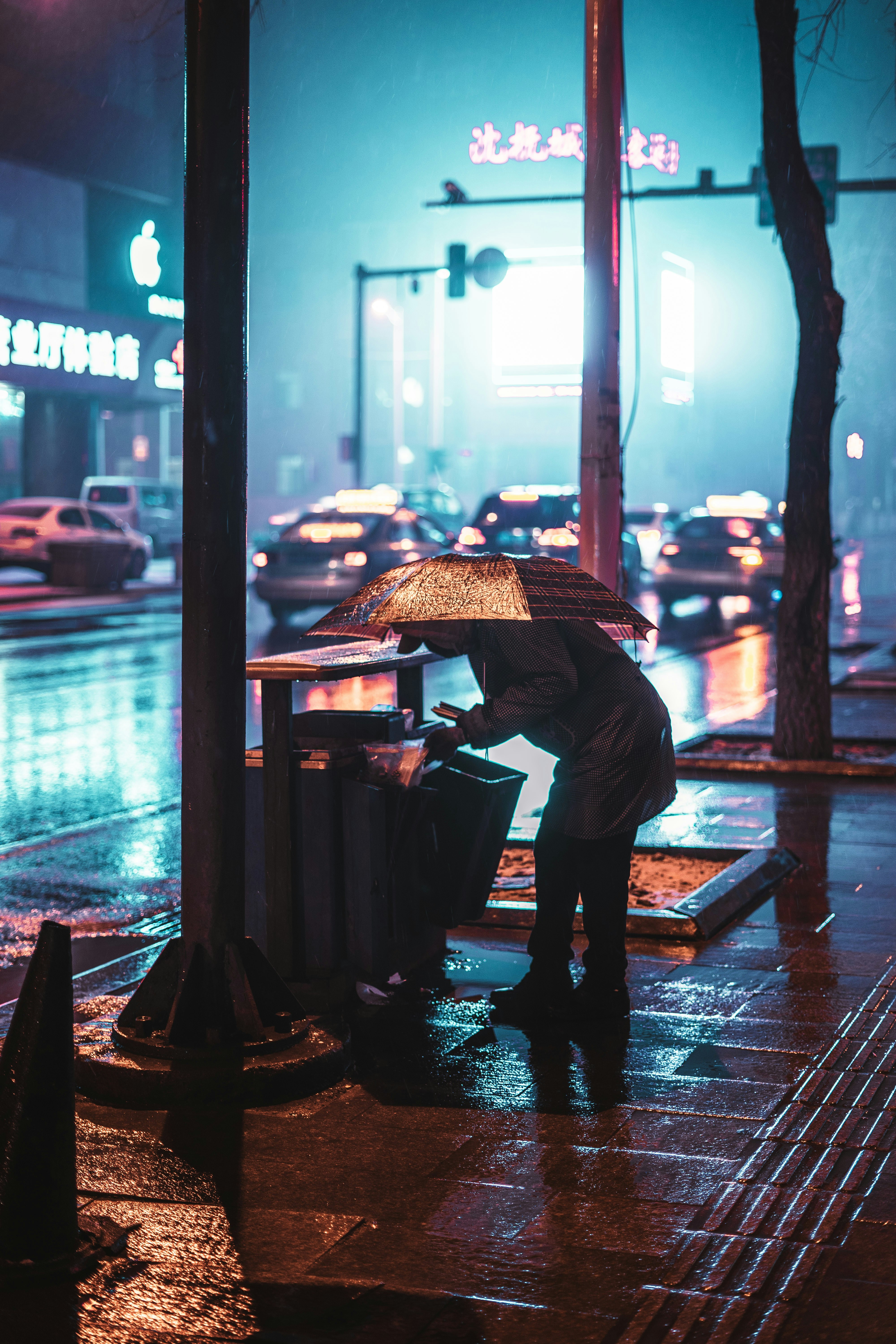 Person with an umbrella searching through a trash bin on a rainy city street at night.