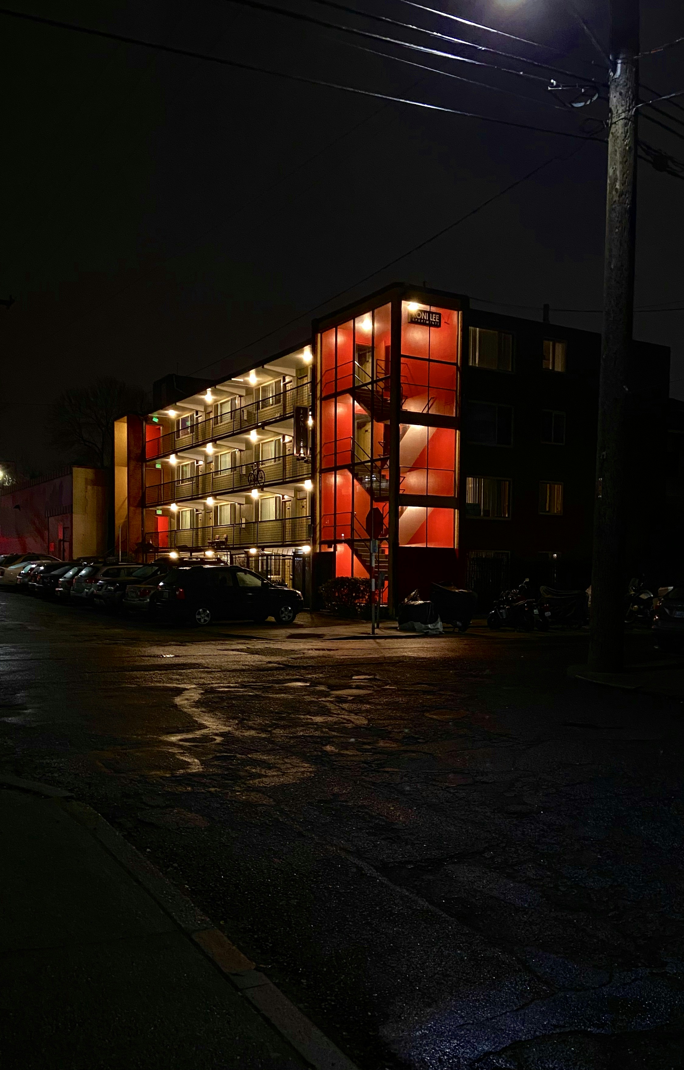 Vibrant red and warm yellow lighting illuminate a multi-story building at night, reflecting off wet pavement. The scene captures an inviting urban atmosphere amidst the darkness.
