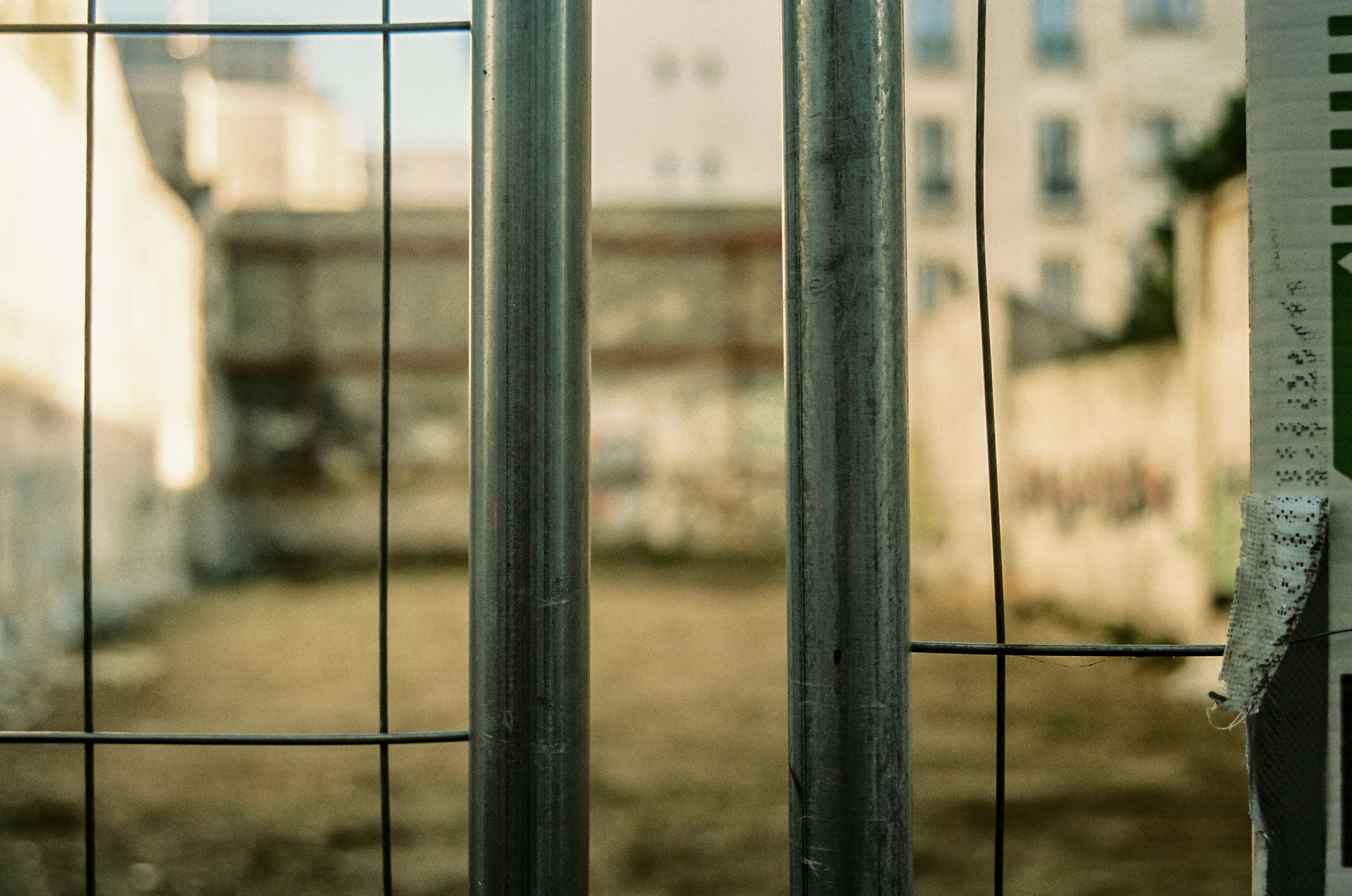 Close-up of vertical metal bars forming a grid, with a blurred urban courtyard beyond. A still-life photograph focusing on industrial textures and shallow depth of field.
