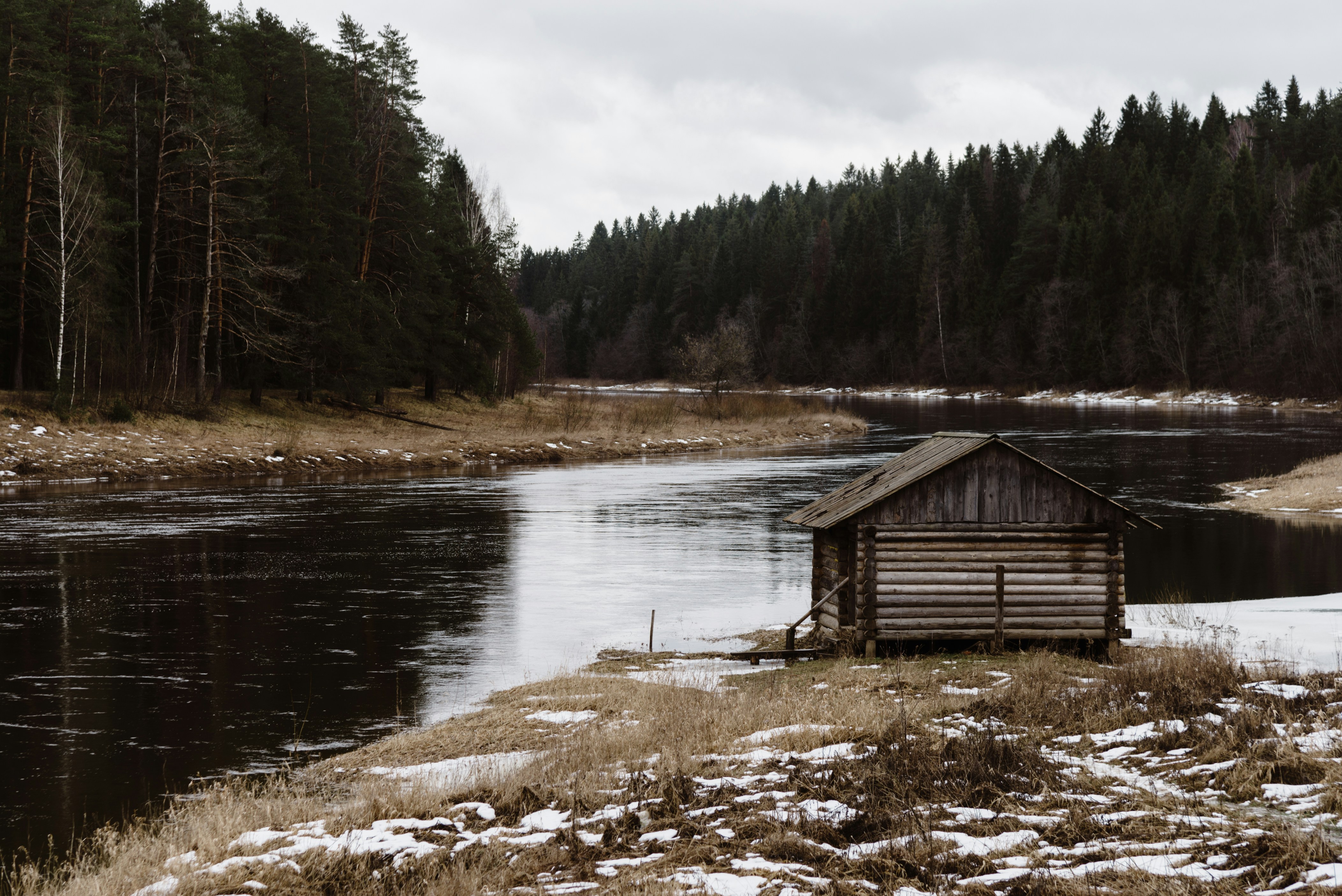 A rustic wooden cabin sits near a partially frozen river, surrounded by sparse grass and patches of snow under a cloudy sky.