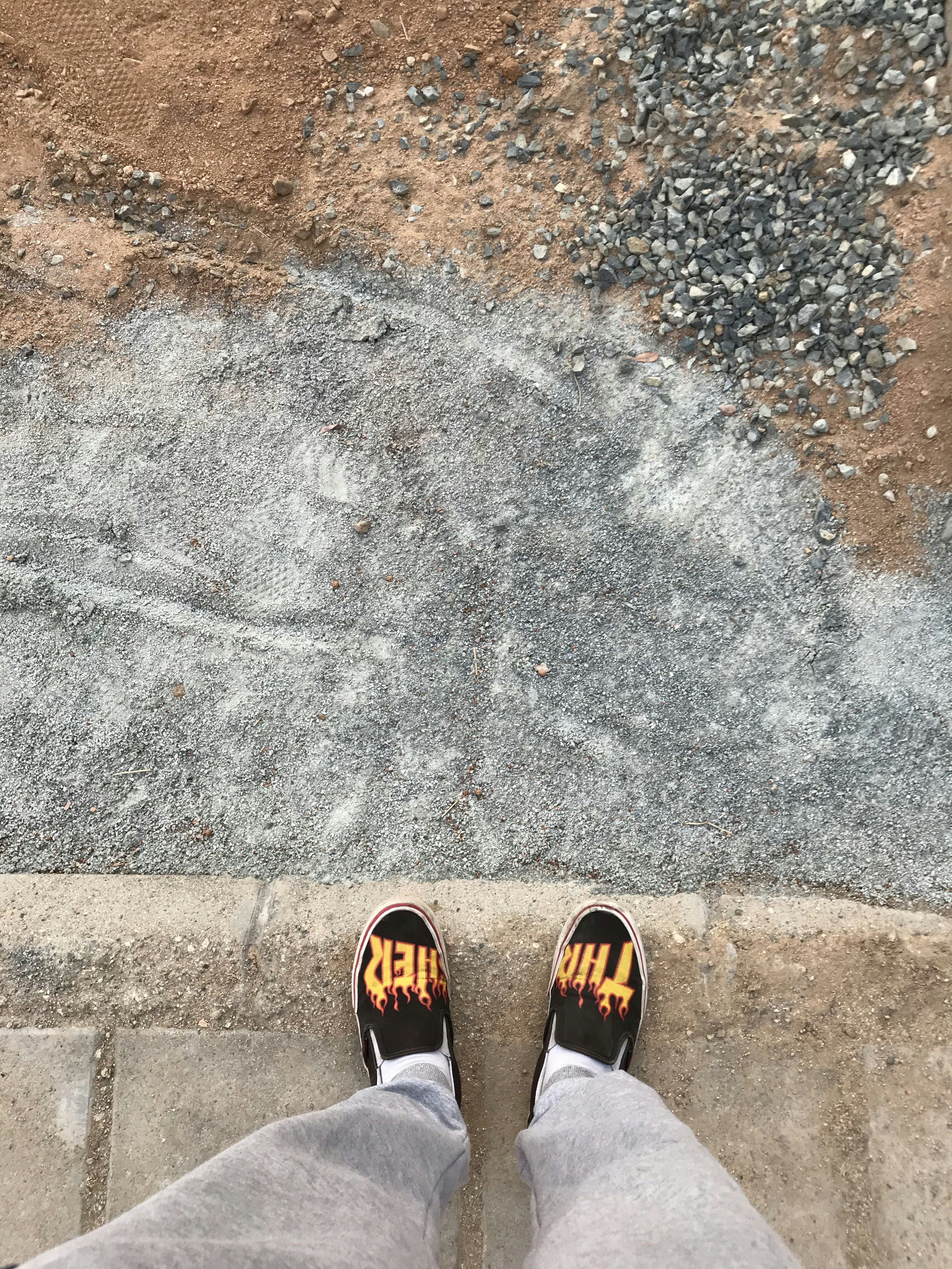 Feet clad in vibrant sneakers stand on a mix of gravel and sand, with a concrete edge visible in the foreground. The image captures a moment of casual style amidst construction materials.