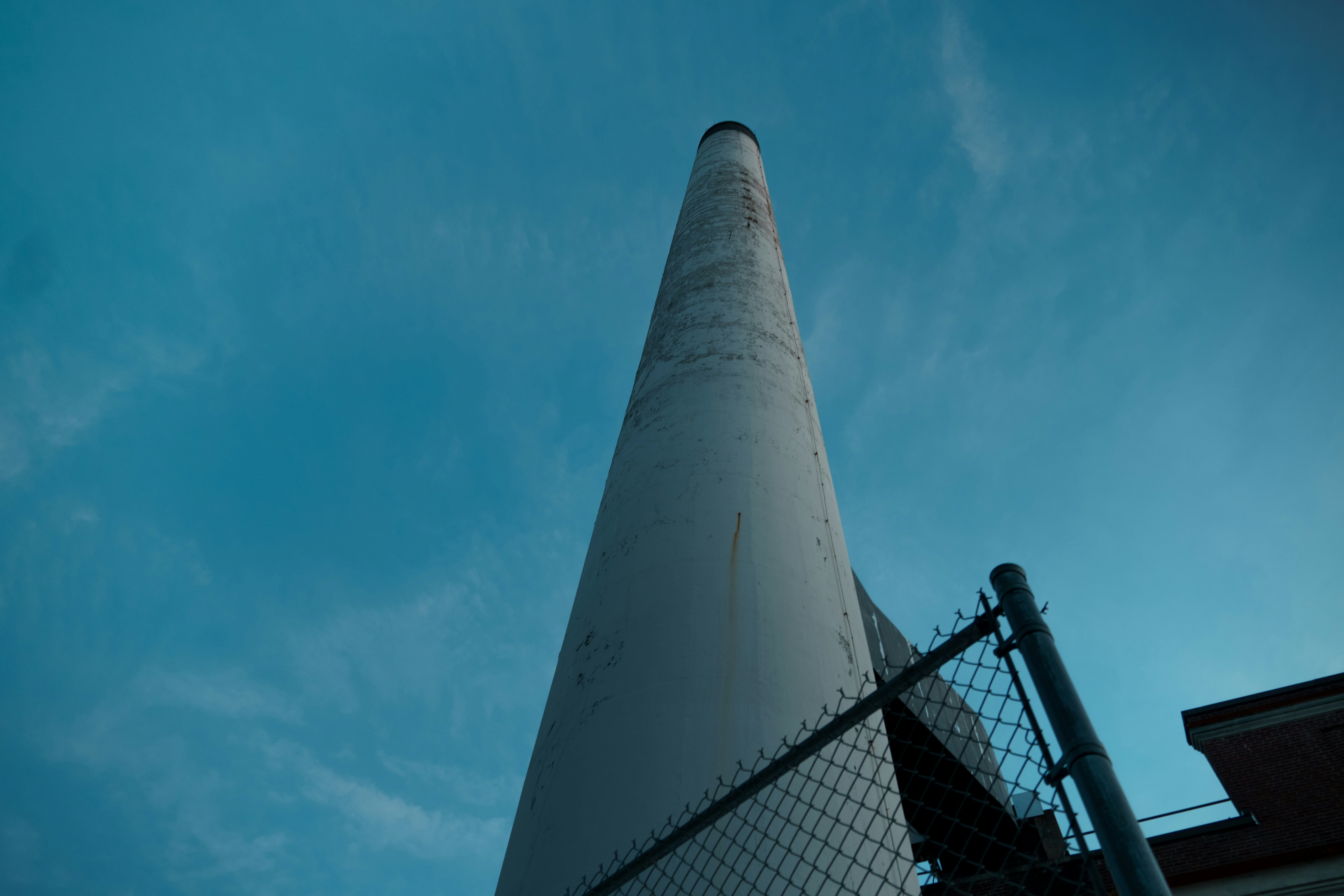 A towering industrial smokestack rises against a backdrop of a vibrant turquoise sky, framed by a chain-link fence.