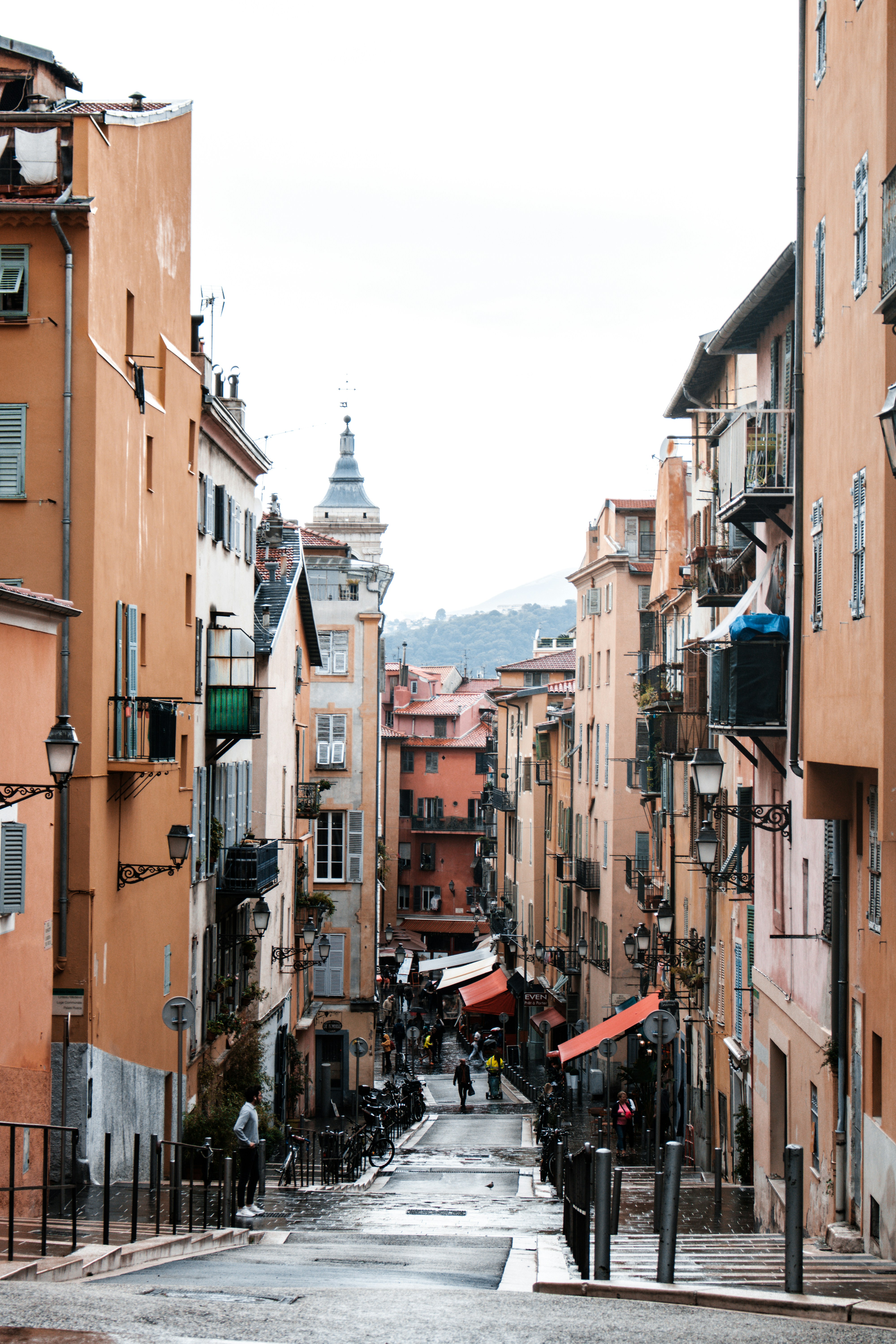 Narrow street lined with colorful buildings leading to a distant clock tower, bustling with pedestrians and bicycles. Rain-soaked pavement reflects the lively atmosphere.