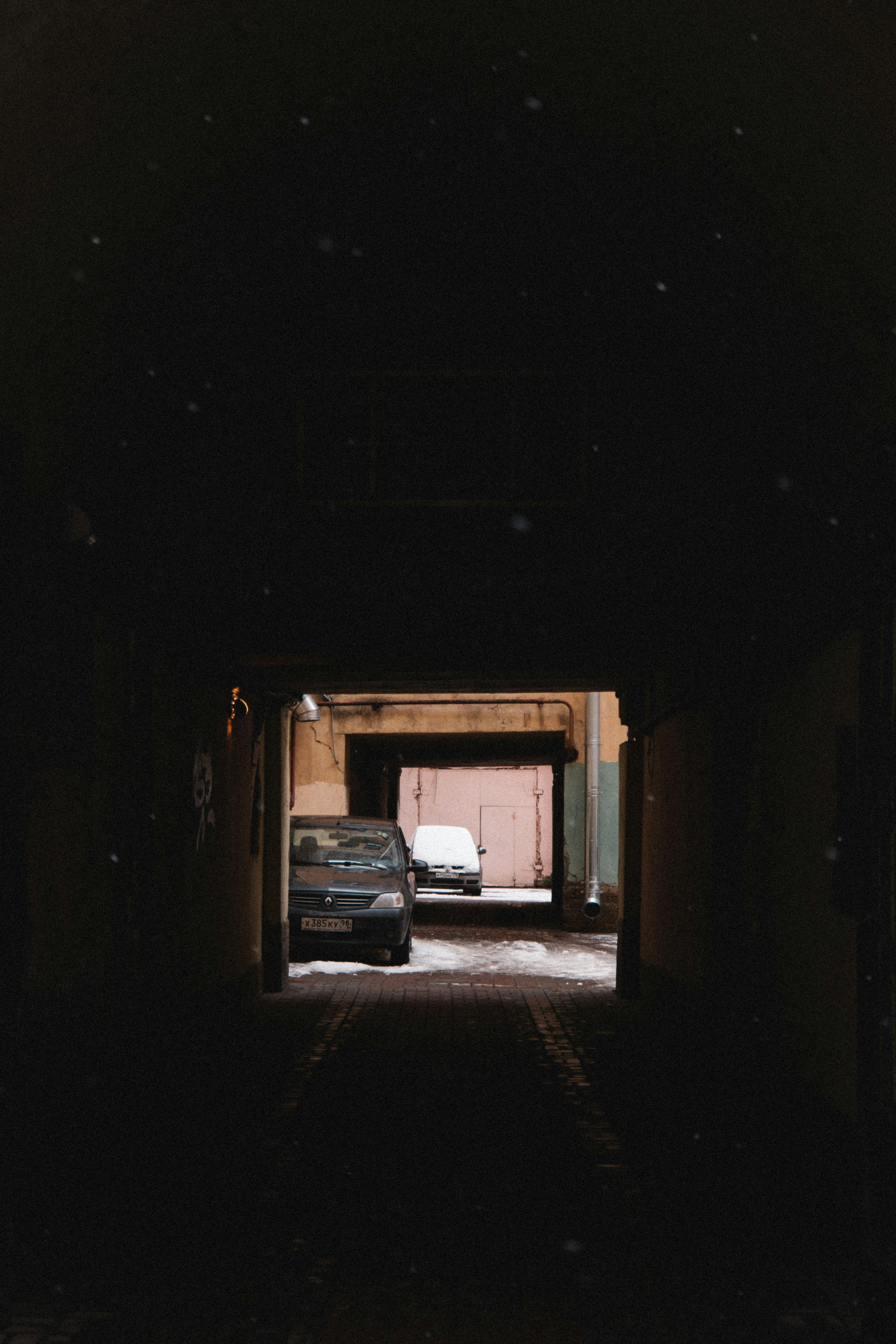 A narrow alleyway reveals parked cars under soft snowfall, leading to an inviting light-filled passageway. The contrast between dark surroundings and illuminated space creates a sense of depth.