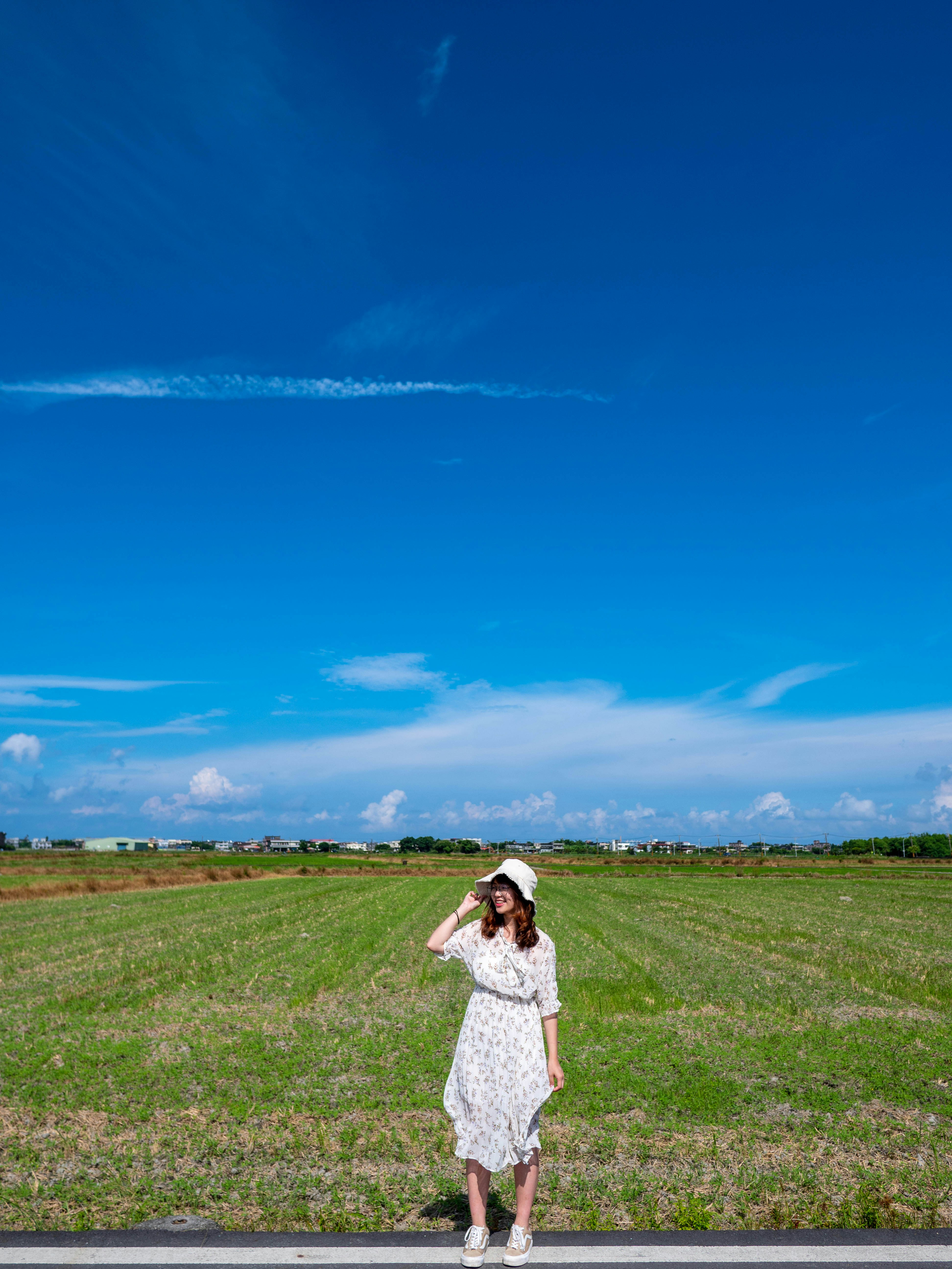 A woman in a white dress and sun hat stands on a road, surrounded by lush green fields under a bright blue sky. The scene captures a moment of tranquility and connection with nature.