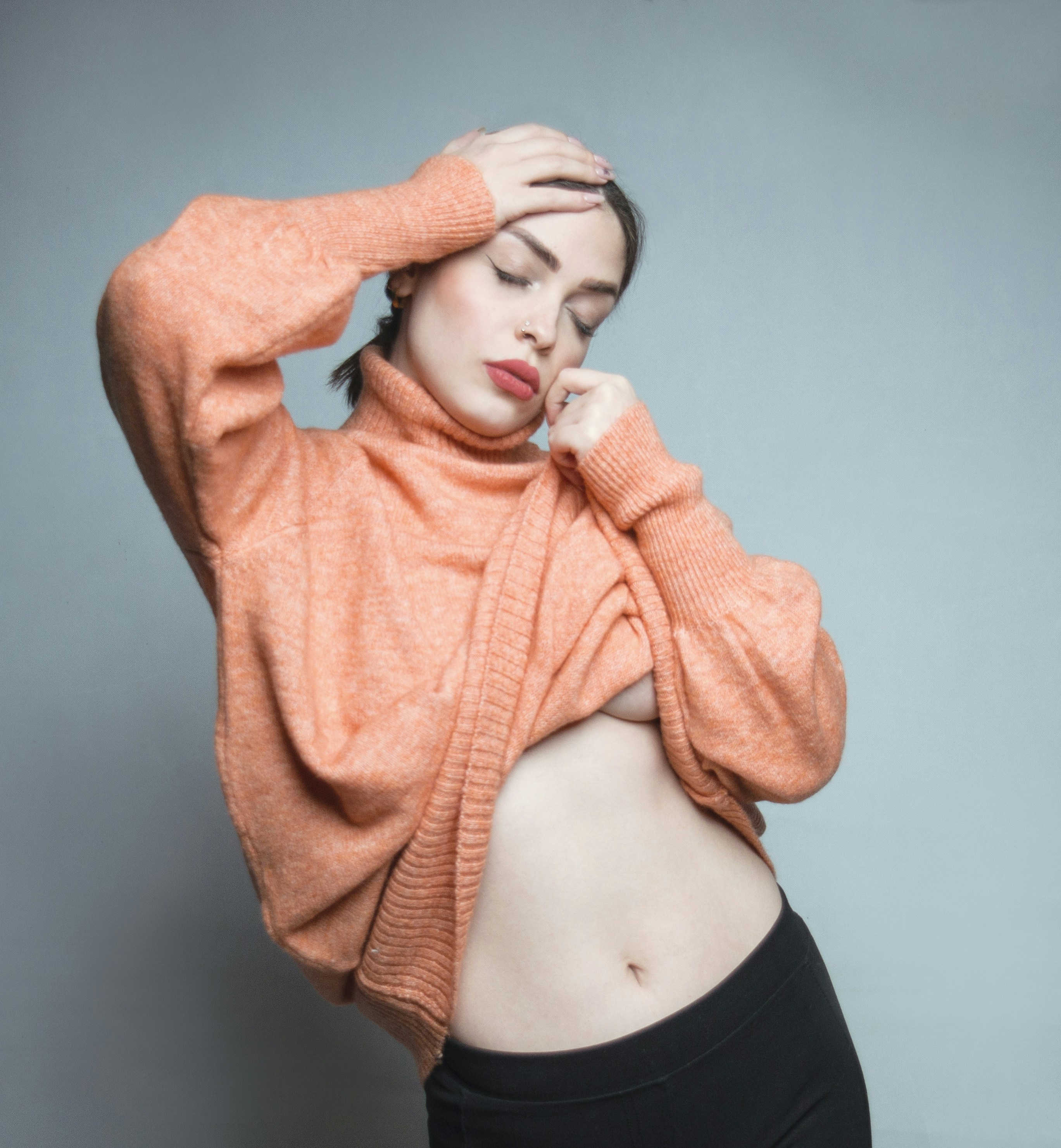 Model in an oversized orange sweater poses with a contemplative expression against a soft gray background.