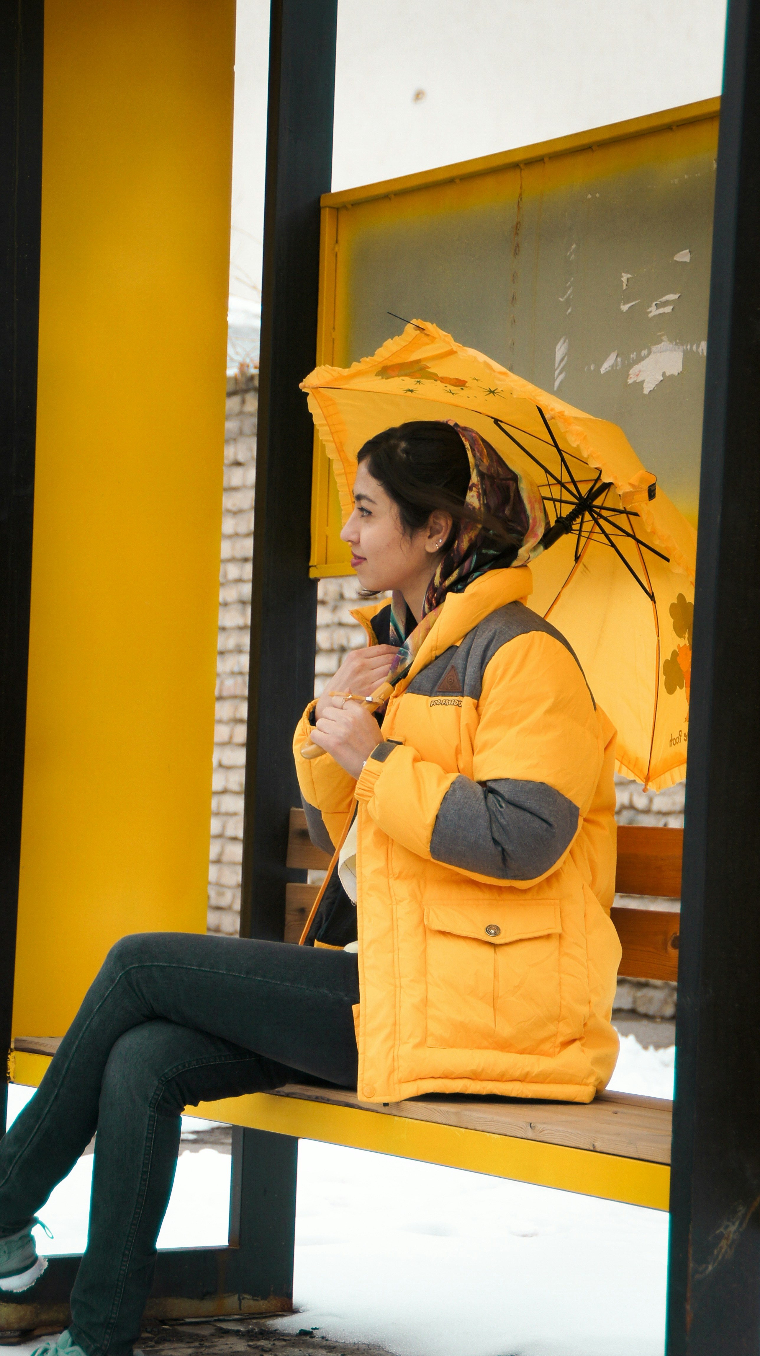 woman in yellow coat and blue denim jeans sitting on yellow concrete bench