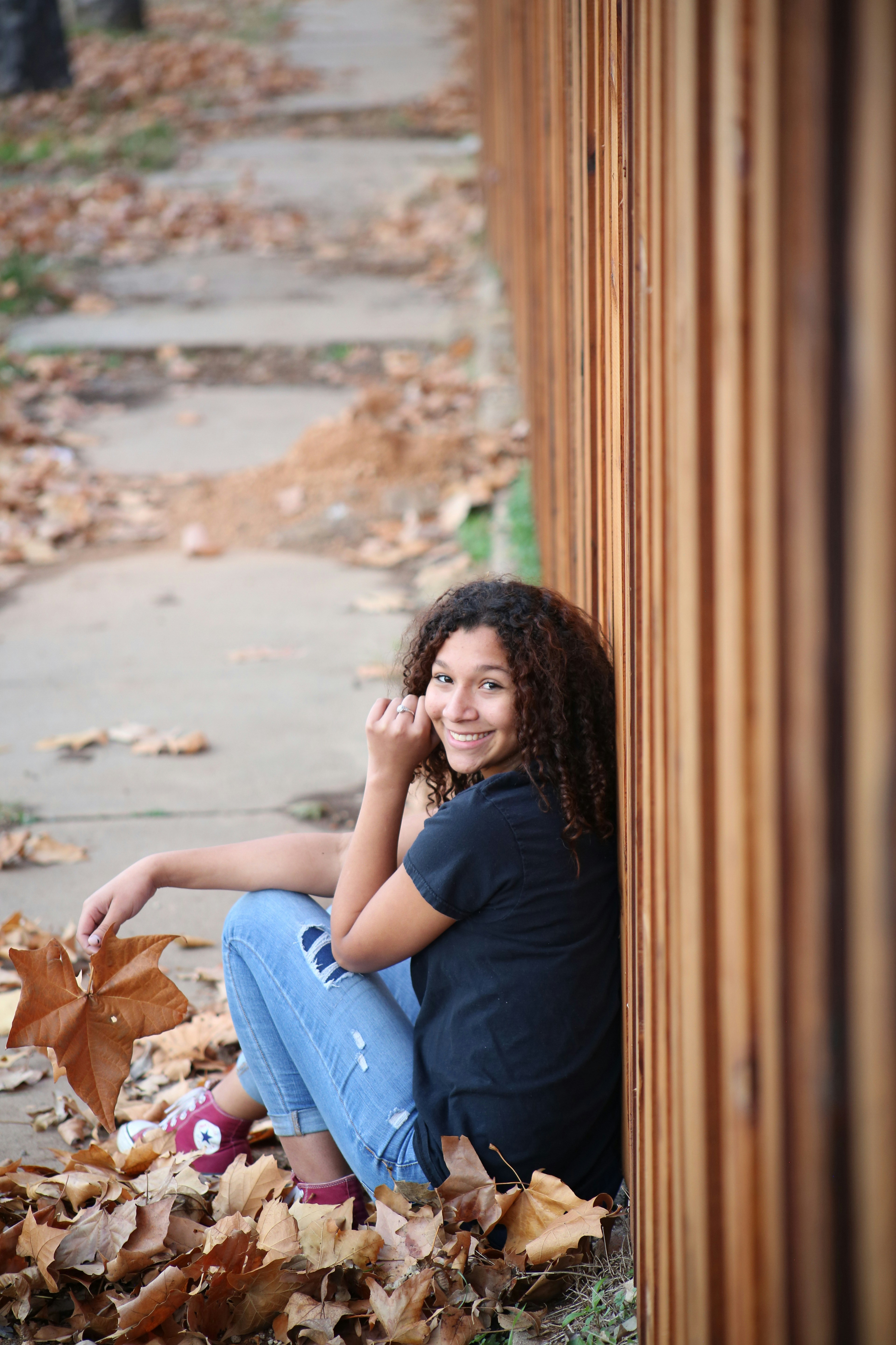 A young girl seated among fallen leaves, playfully holding a large leaf while smiling at the camera. The wooden fence in the background adds warmth to the scene.