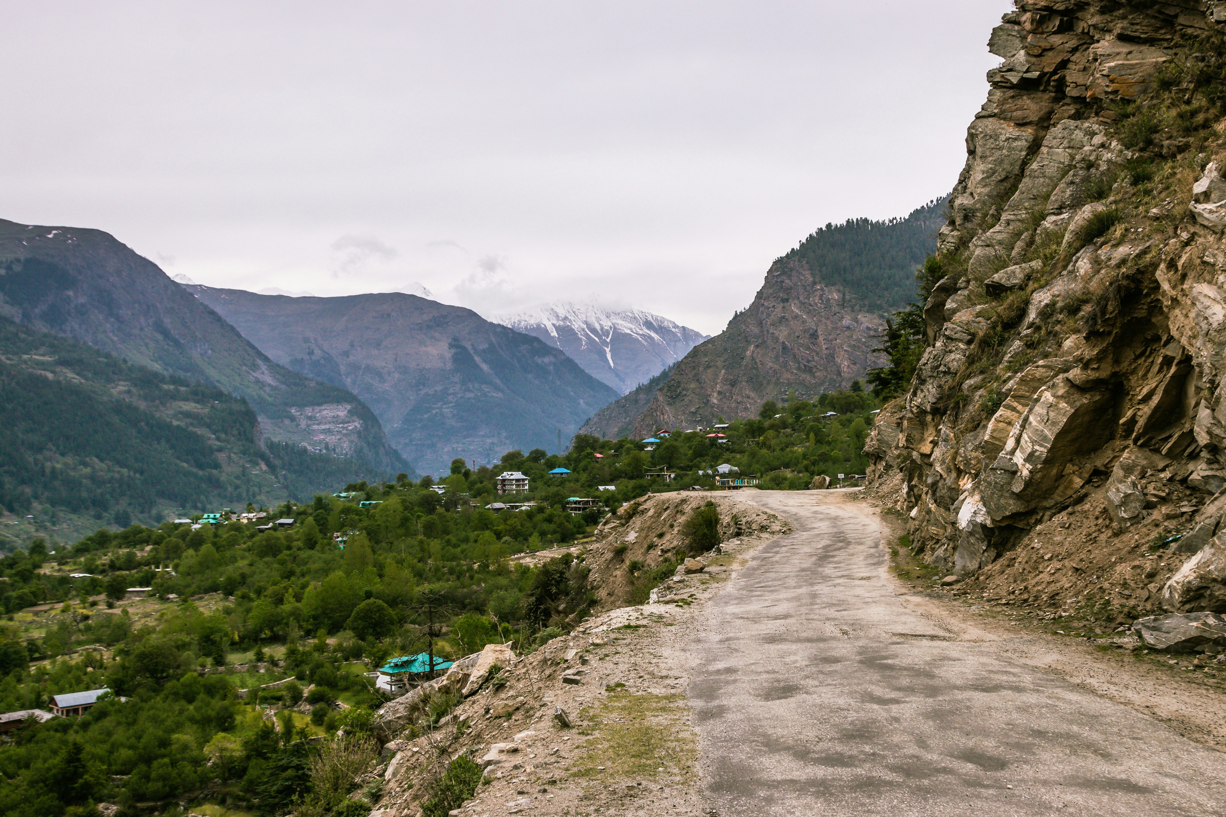 A narrow road curves along a rocky hillside, surrounded by lush greenery and distant snow-capped peaks, showcasing the beauty of mountainous terrain.