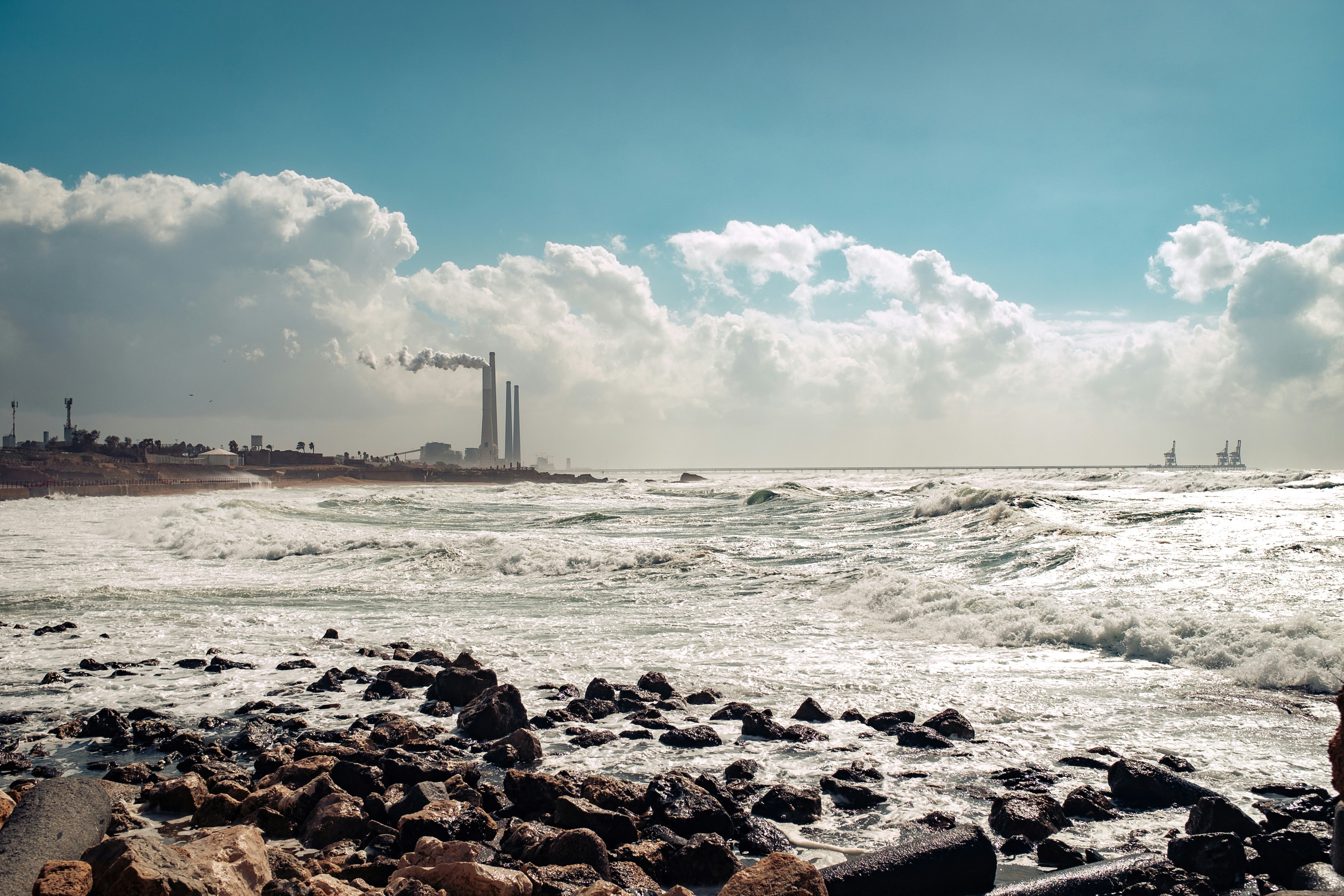 Waves crashing against rocky shore with industrial smokestacks in the background under a cloudy sky.