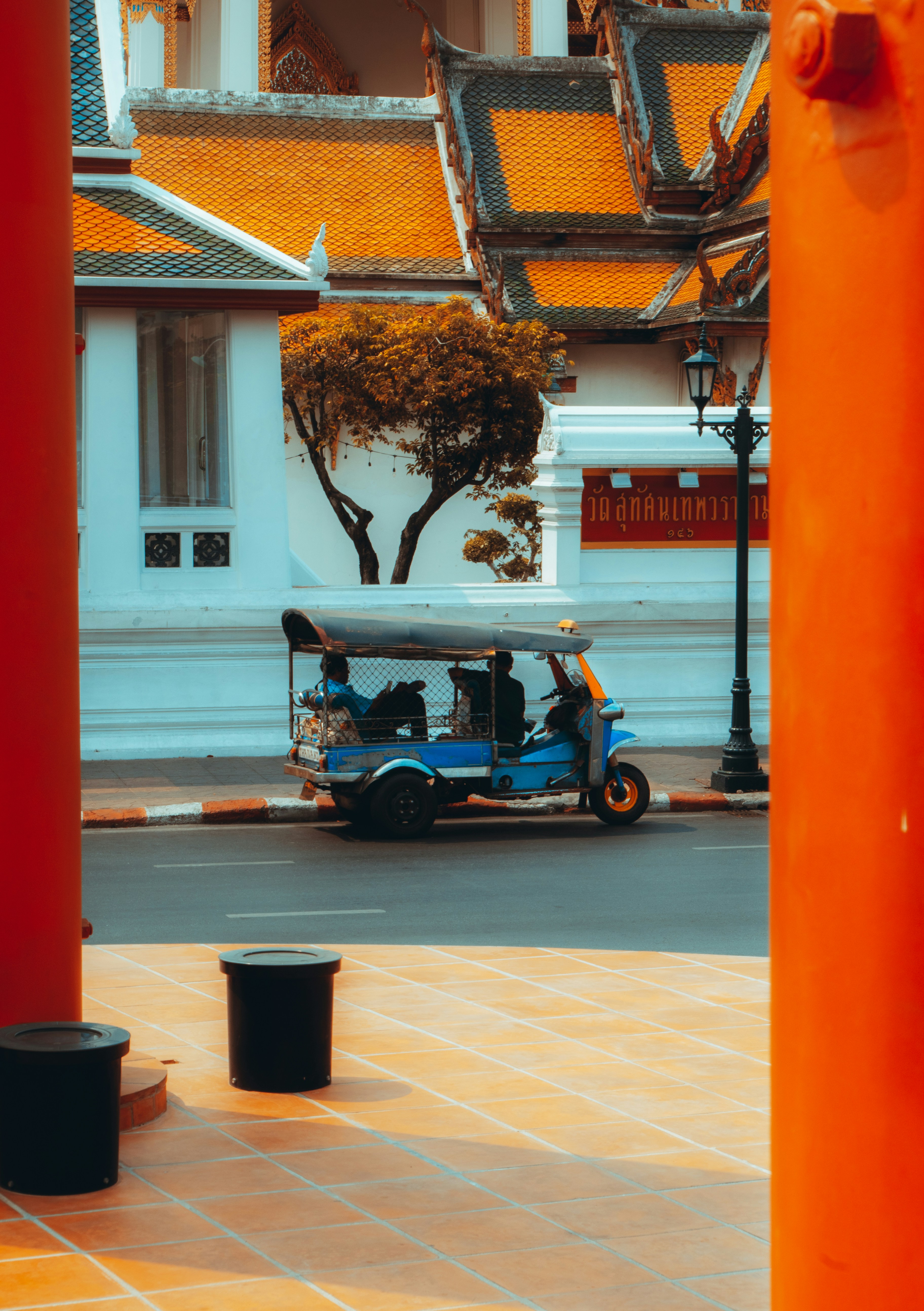 A vibrant blue tuk-tuk parked beside a white building with striking orange roofs, framed by red pillars. The scene captures the essence of urban life and cultural architecture.