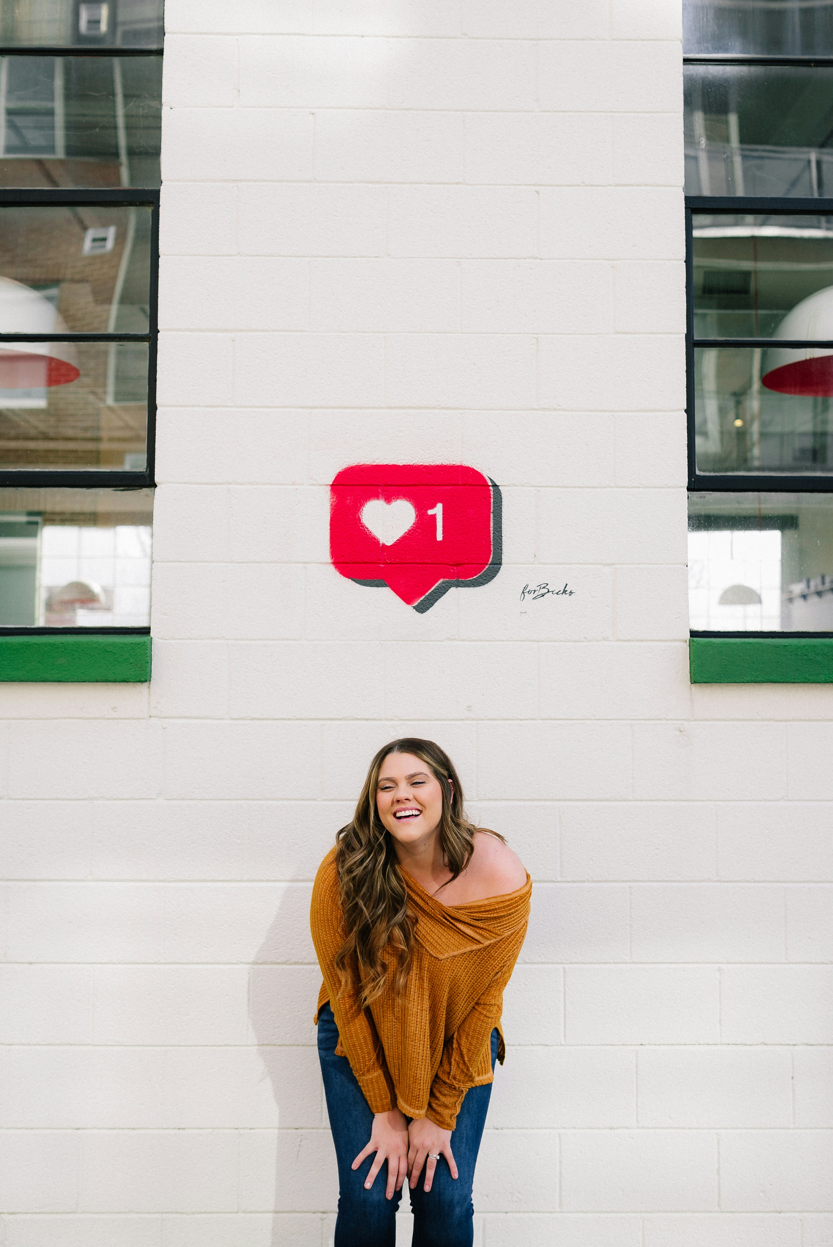 Woman in brown sweater laughing beneath a painted social media like symbol on a white brick wall.