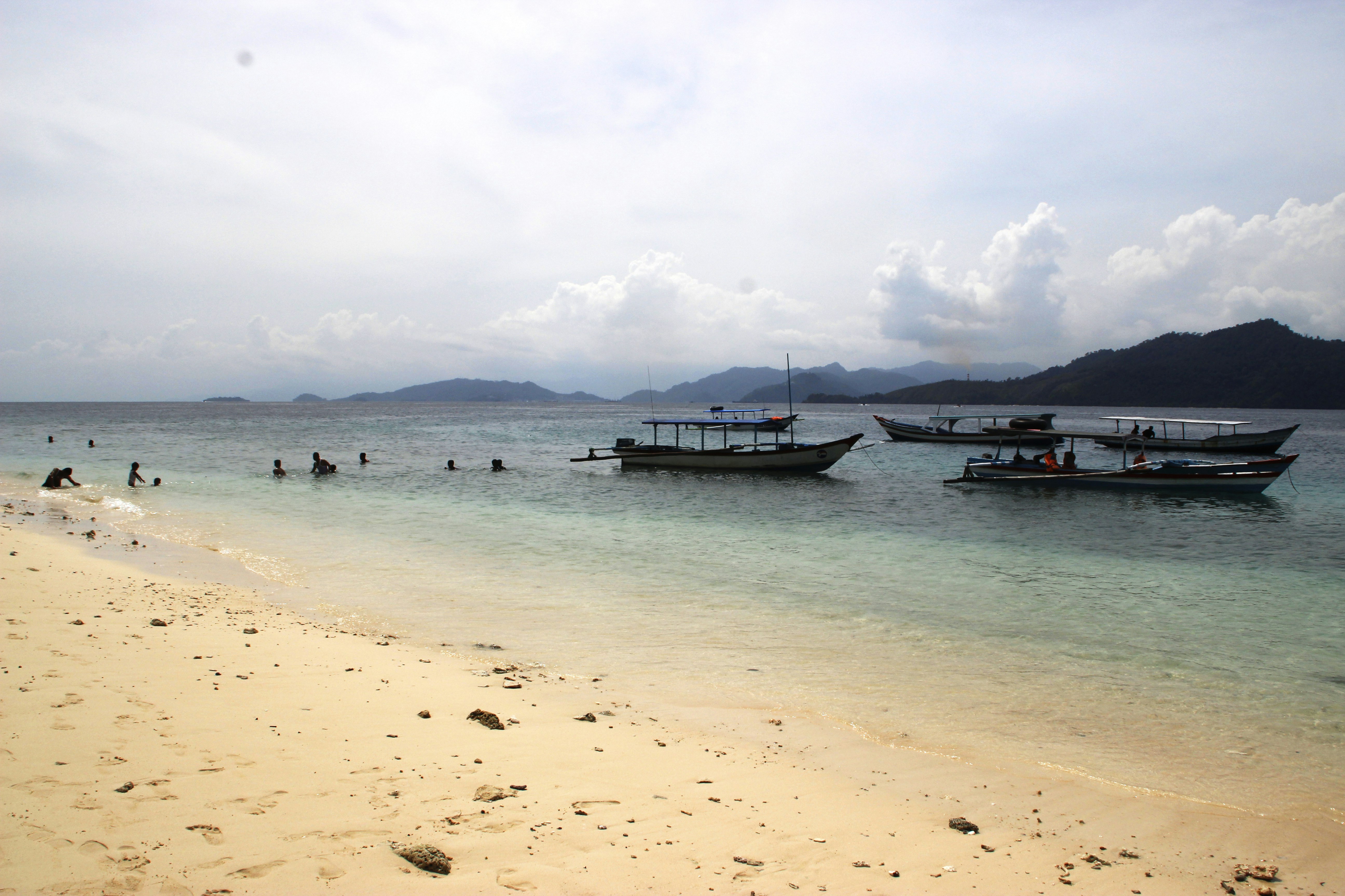 Sandy beach leading to calm turquoise waters with anchored boats under a cloud-streaked sky.