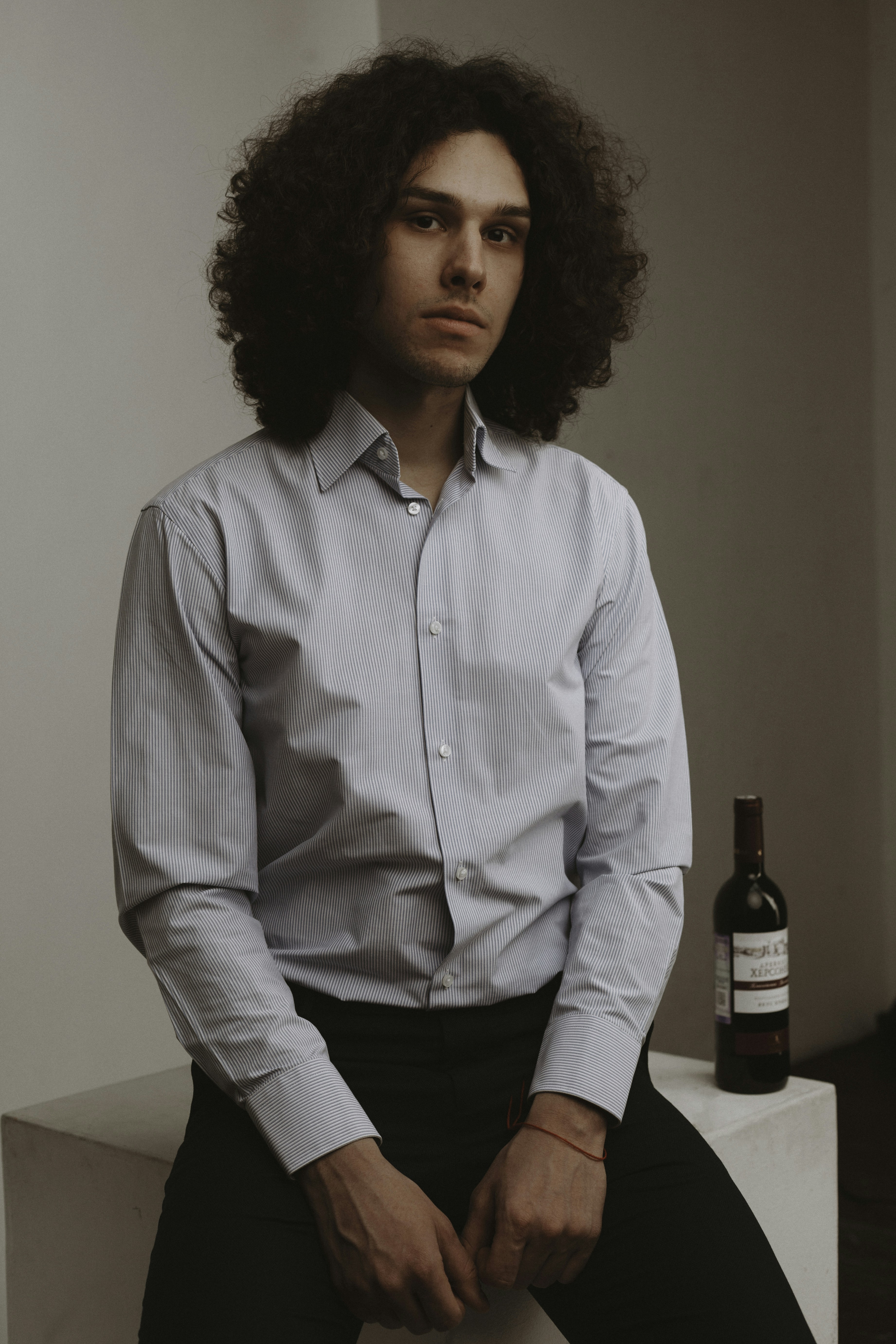 Young man in a striped shirt seated on a pedestal, with a bottle of red wine beside him. The atmosphere is calm and introspective.