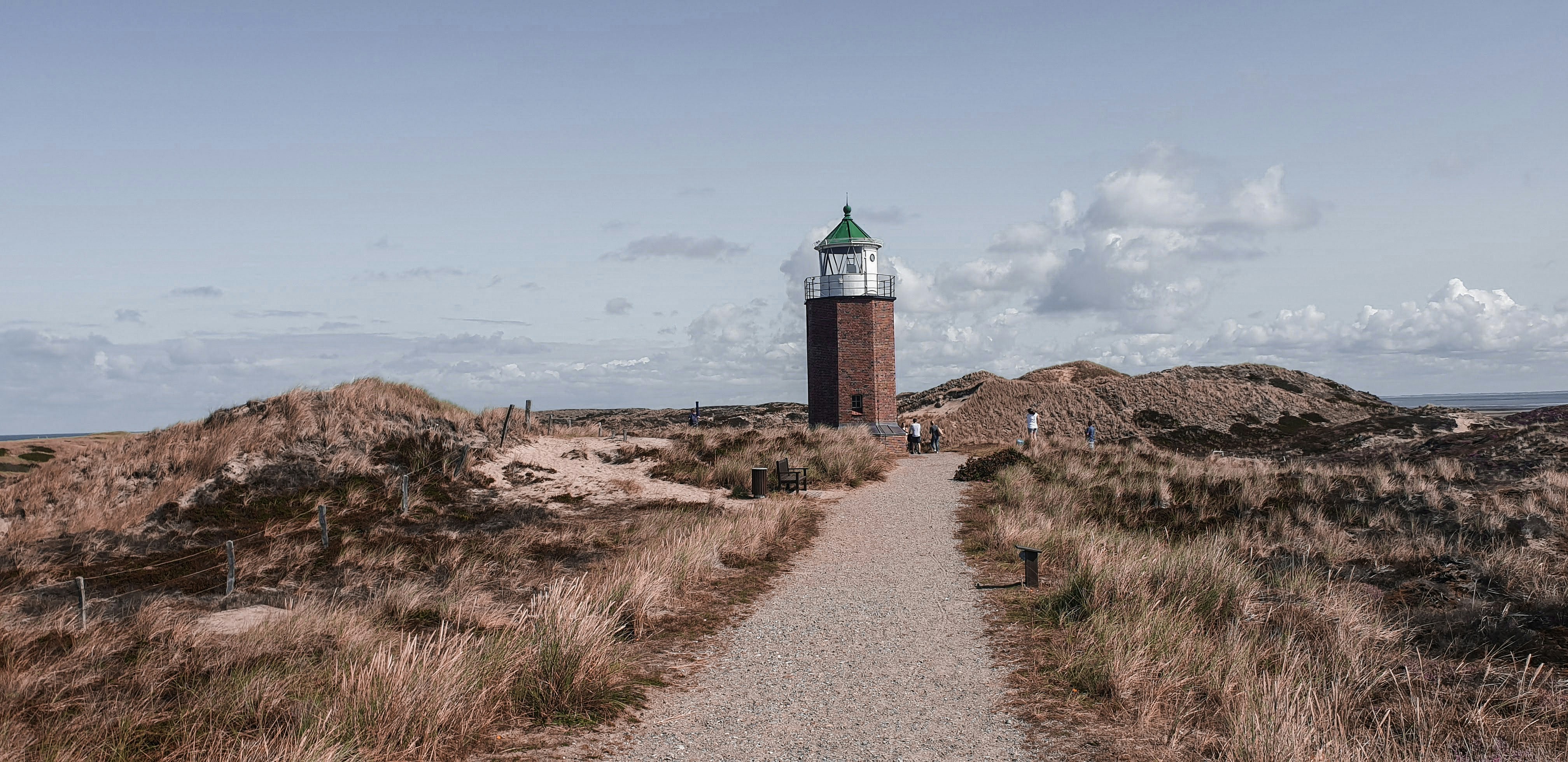 Lighthouse stands solitary on a coastal path flanked by sandy dunes under a partly cloudy sky.