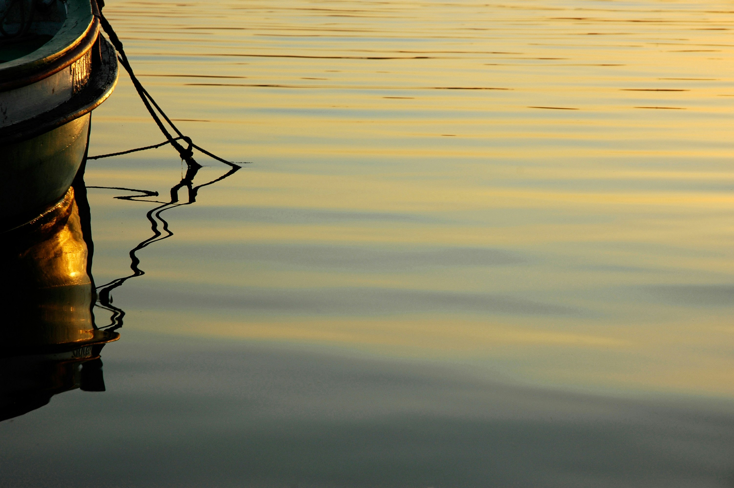 Boat hull and mooring rope reflected on calm water at golden hour, with soft ripples and warm sunset hues.