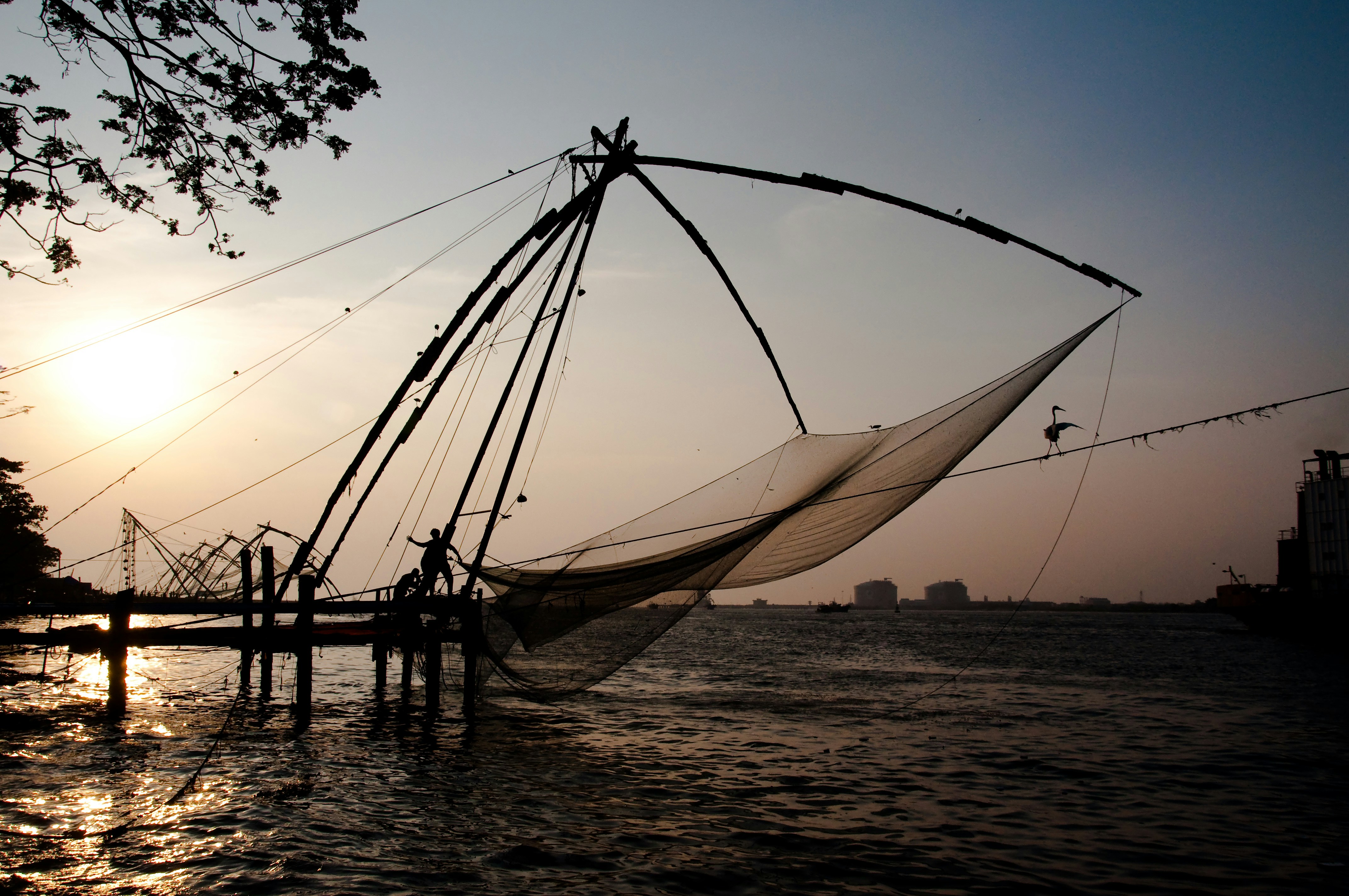 Silhouetted Chinese fishing nets against a sunset sky over a tranquil waterway.