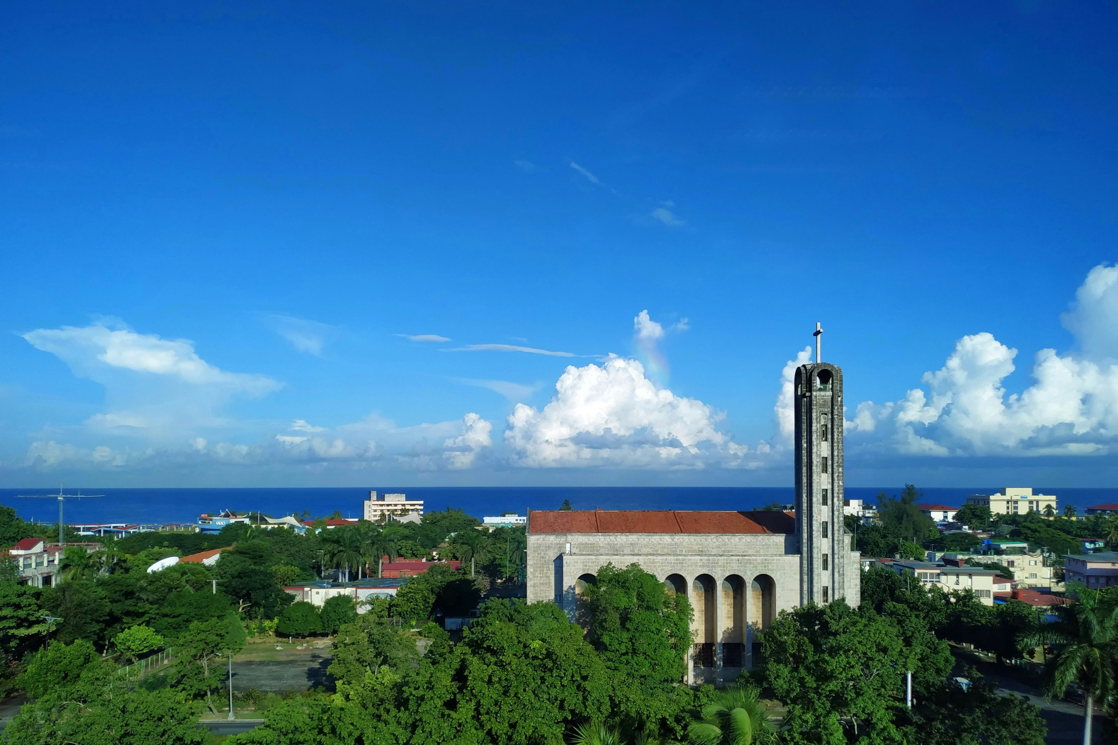 Church tower rising above lush greenery against a backdrop of ocean and dramatic clouds.