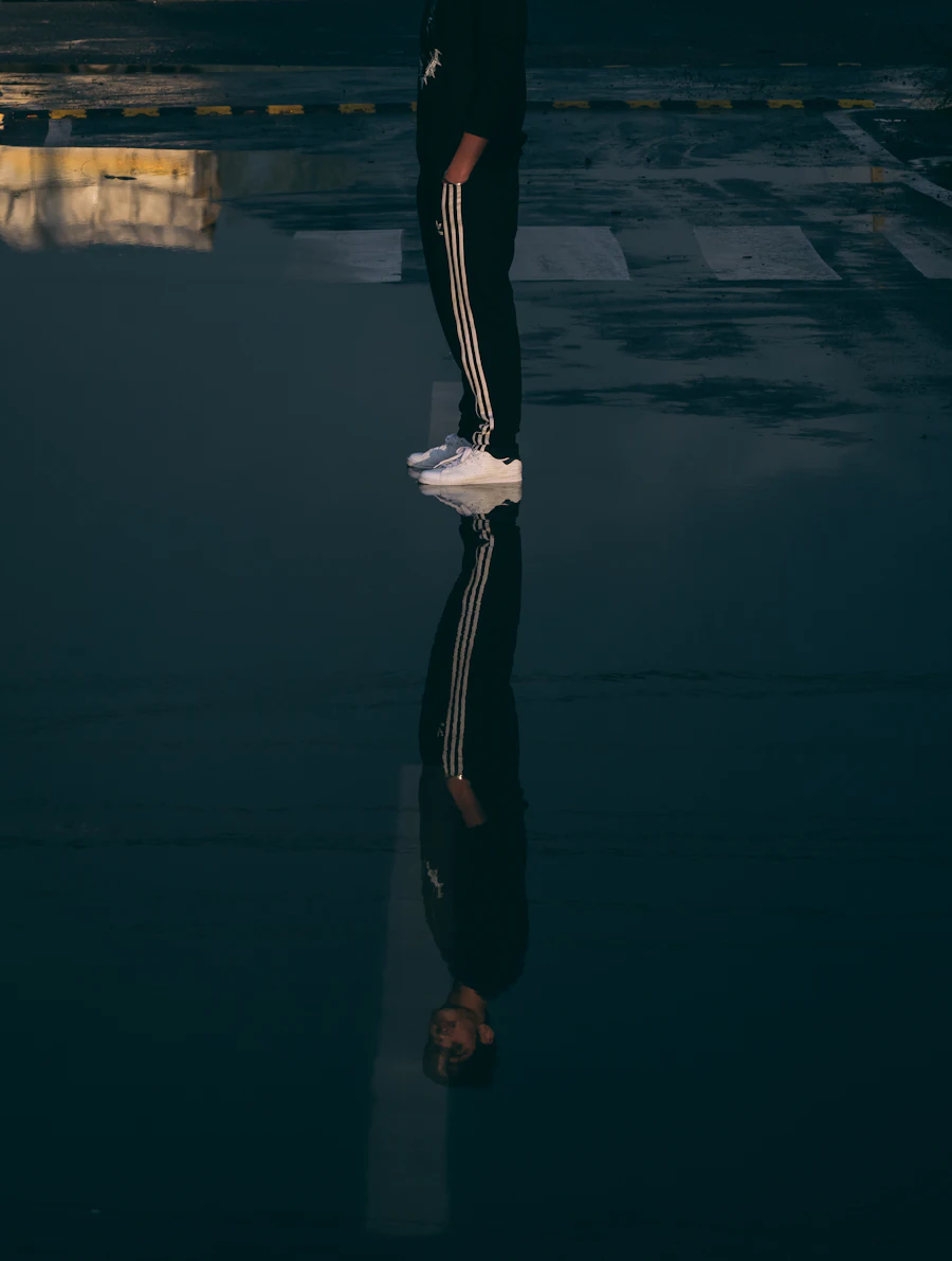 A lone figure stands on a wet surface, their reflection mirrored in the puddle below, creating a striking visual interplay of reality and illusion.