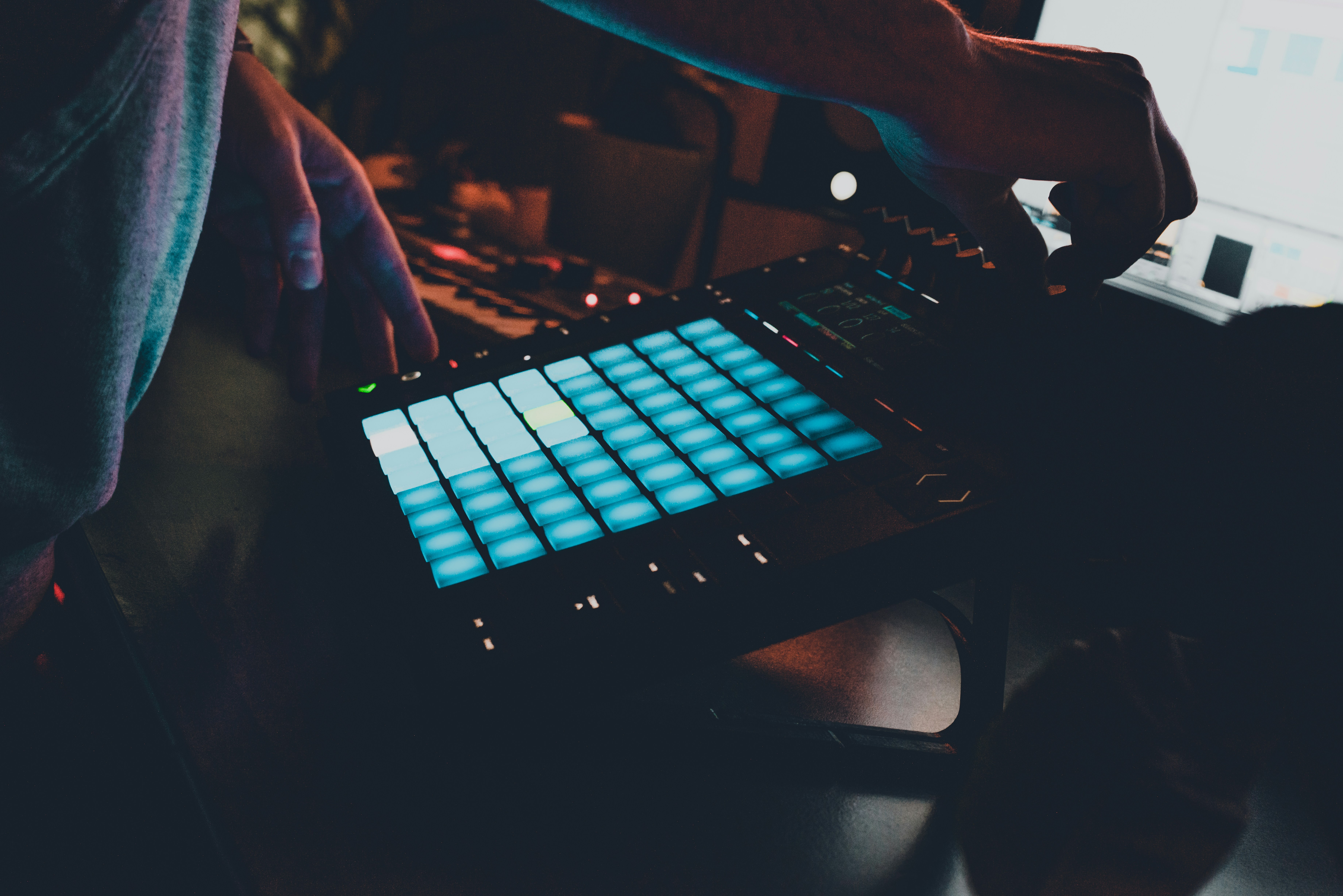 Close-up of a musician's hands on a digital music pad, illuminated by vibrant blue lights, as they create electronic beats in a studio setting.