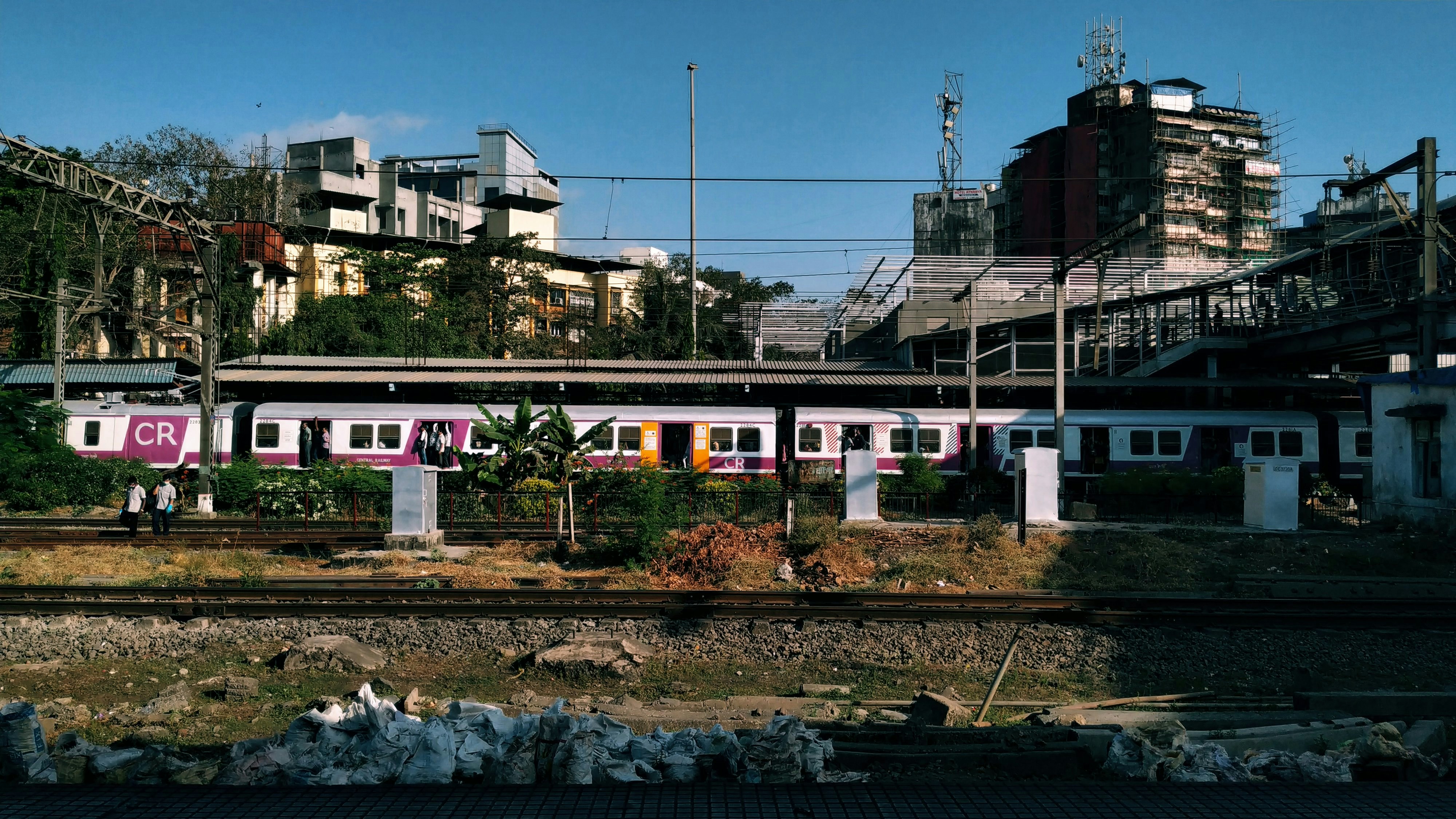 Row of purple-and-white commuter cars on elevated tracks beside an urban platform, with city buildings and a clear blue sky in the background.