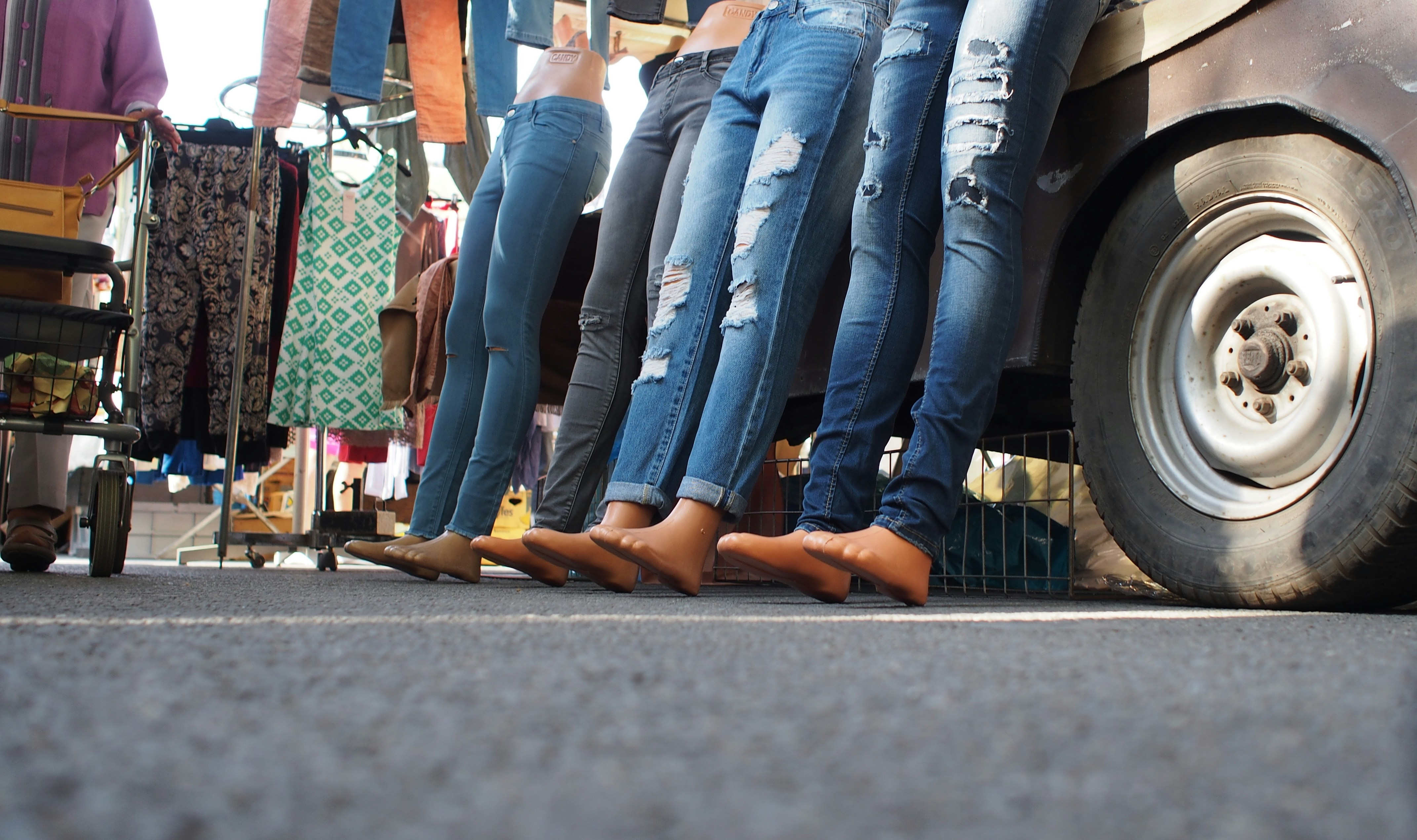 Low-angle street photograph focusing on a row of denim-clad legs and brown boots beside a market stall and a vehicle, capturing everyday urban commerce.