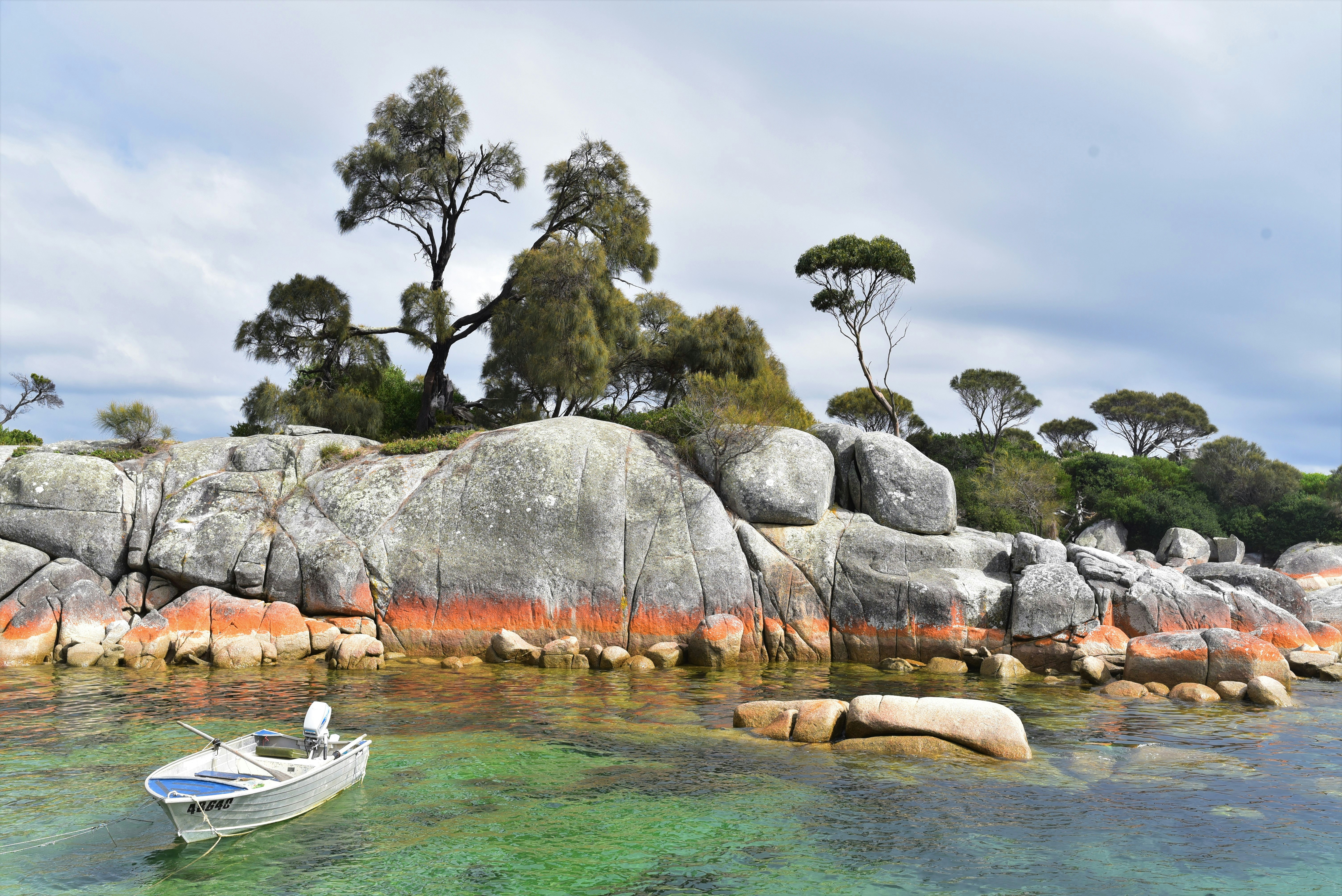 A small boat gently floats in clear waters beside rugged rocks and scattered trees, showcasing a tranquil coastal landscape.