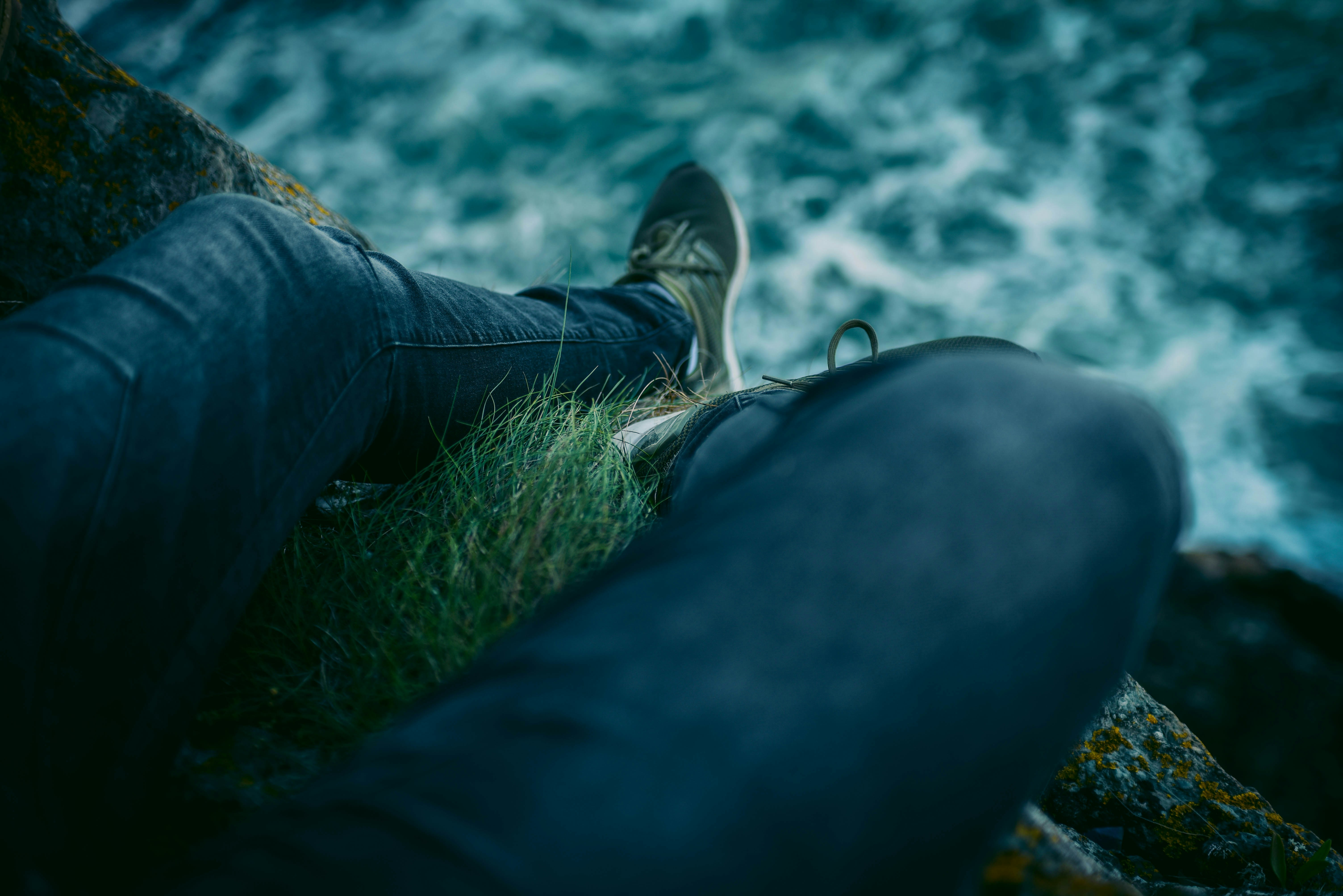 A person’s legs dangle over a rocky cliff, surrounded by vibrant green grass, with crashing waves below. The scene captures a sense of adventure and contemplation.