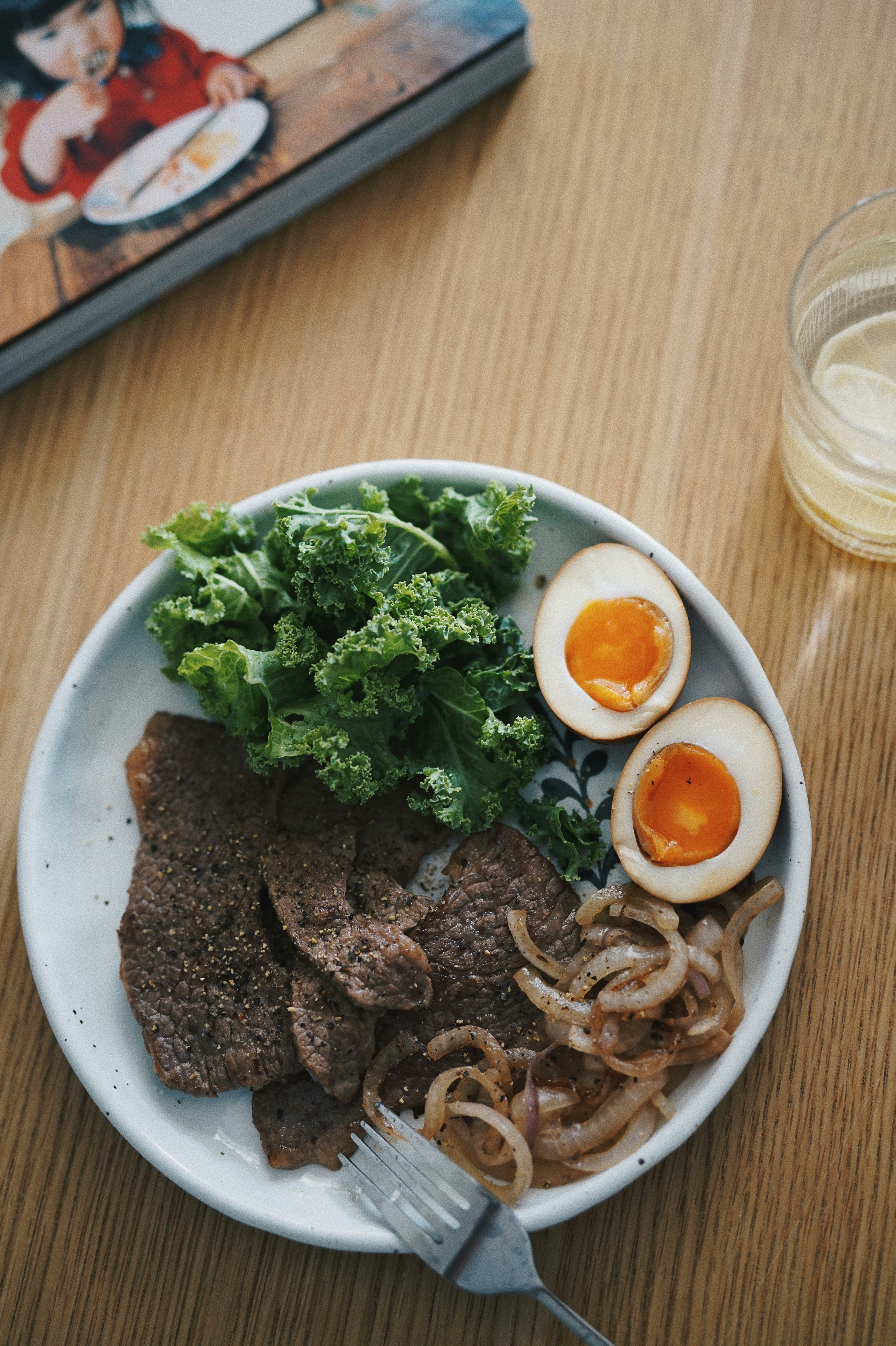 Tray of savory beef, soft-boiled eggs, and kale with a lemon-glass on a wooden table