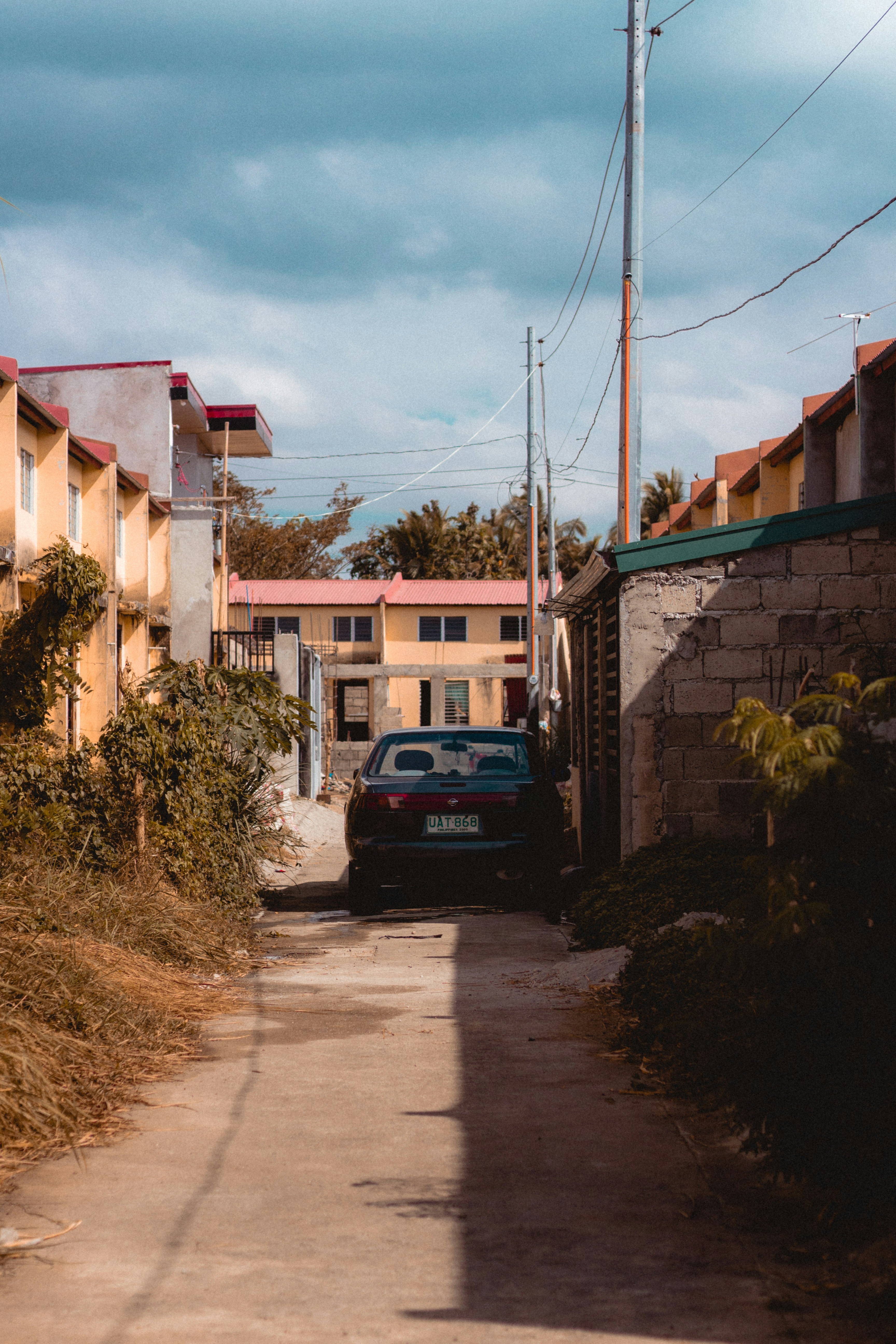 Narrow alleyway flanked by residential buildings, featuring a parked car and overgrown vegetation. The scene captures a moment of urban tranquility.