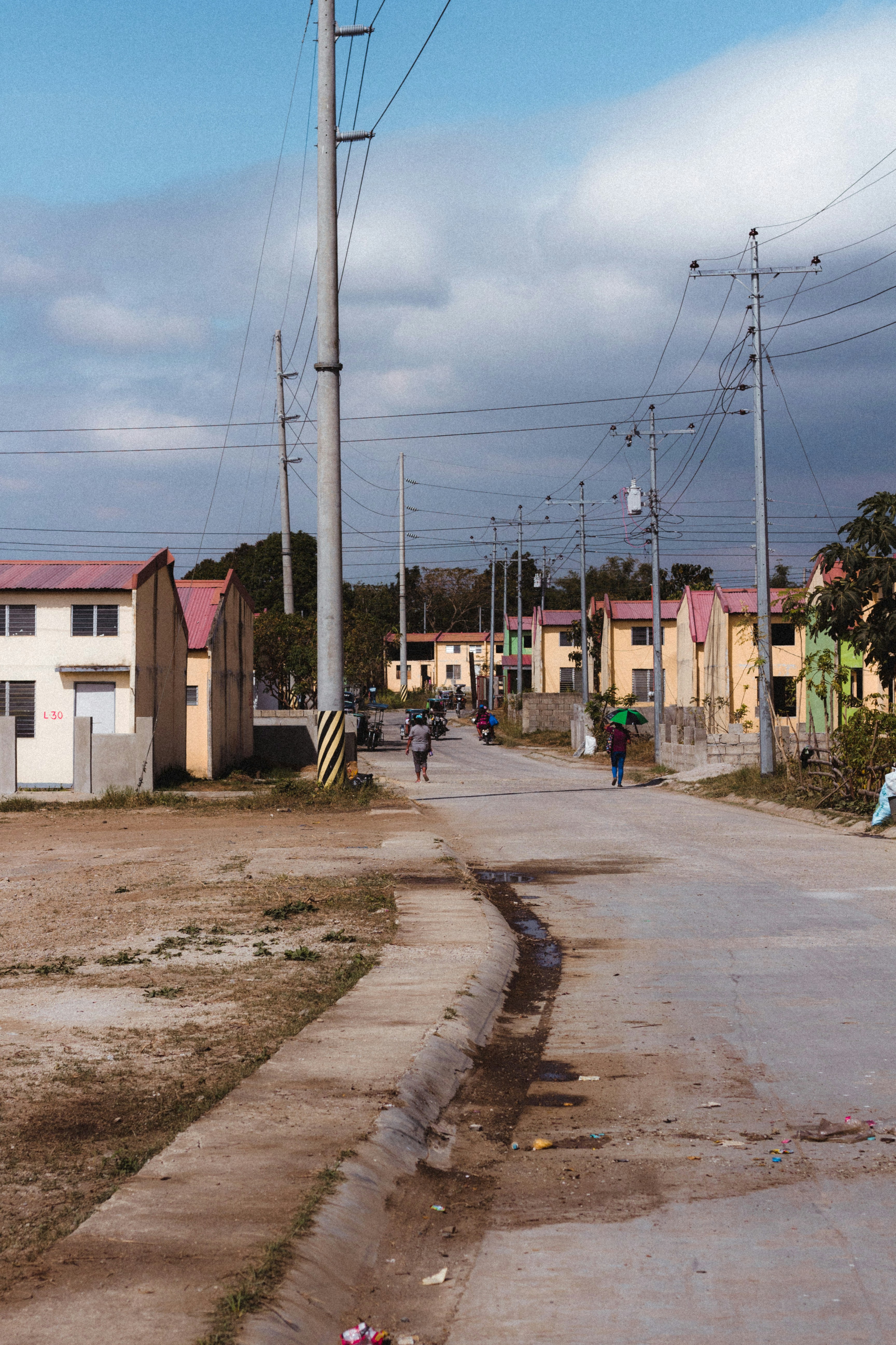 Residential street lined with colorful houses and utility poles under a partly cloudy sky. People walk along the road, adding life to the scene.